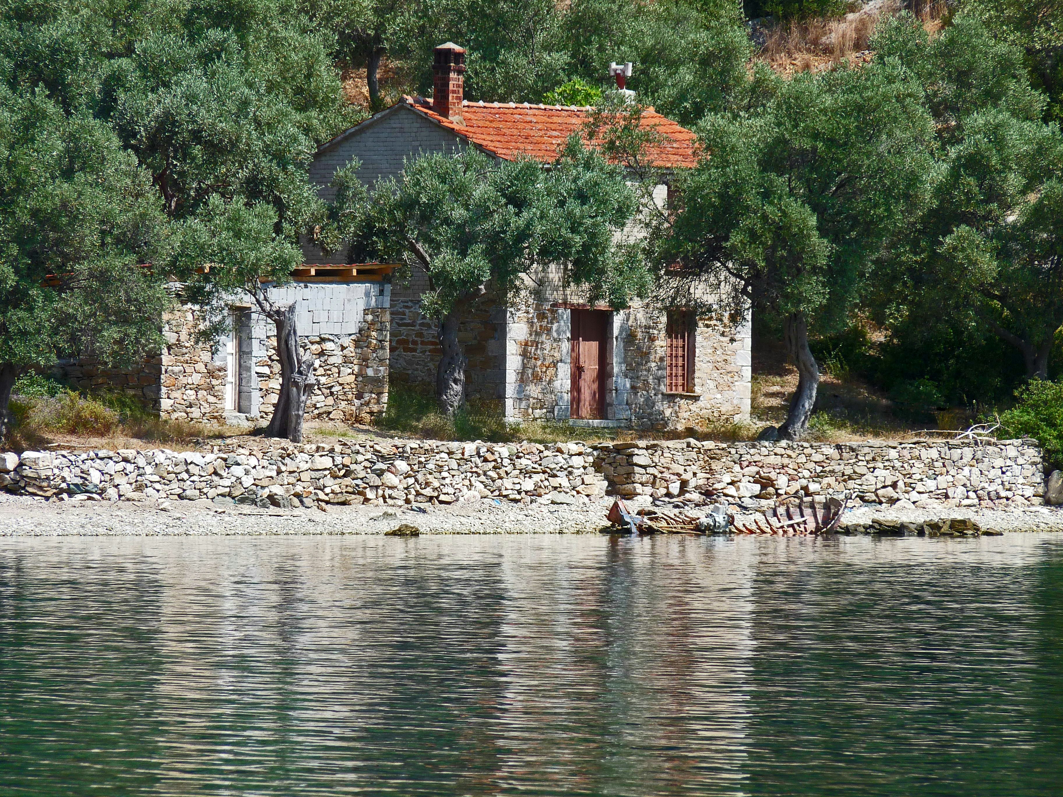 Stone cottage with a red-tiled roof sits along a rocky lakeshore framed by olive trees. A small boat rests at the water's edge, its reflection rippling in the calm surface.