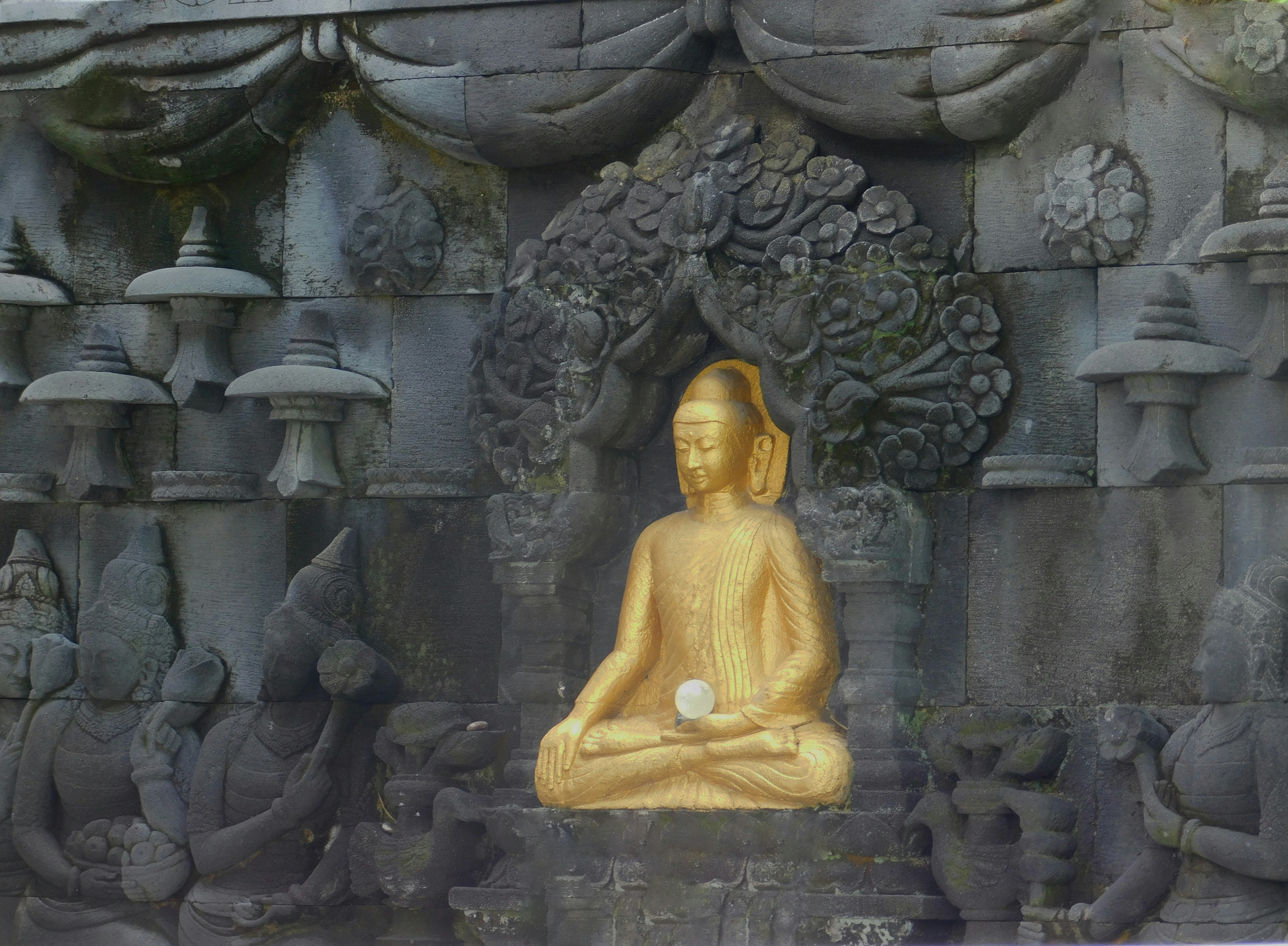 Golden statue of Buddha seated in meditation, surrounded by intricately carved stone figures and floral designs on a temple wall.