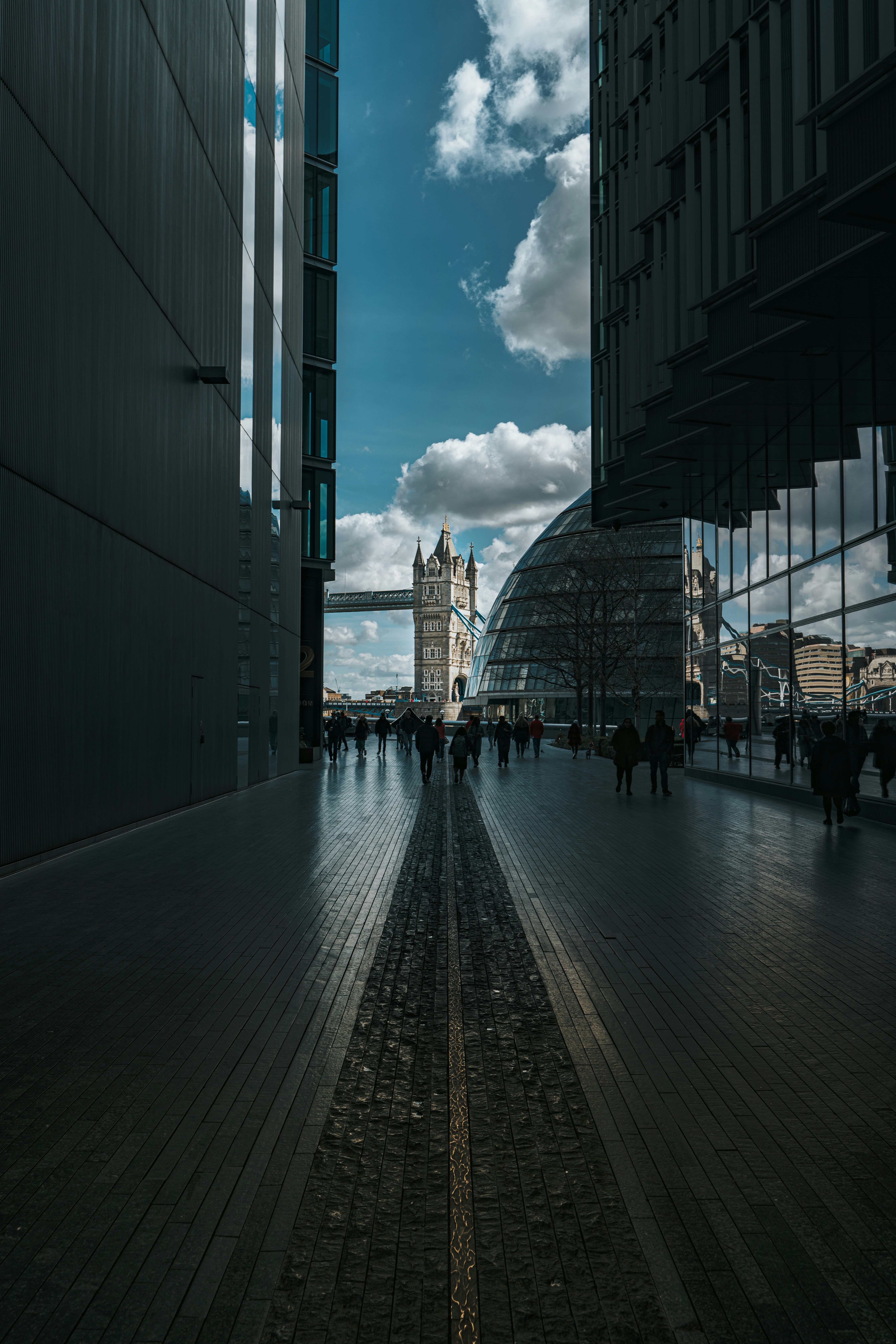 A modern urban pathway framed by sleek buildings, leading to the iconic Tower Bridge under a dramatic sky filled with clouds.