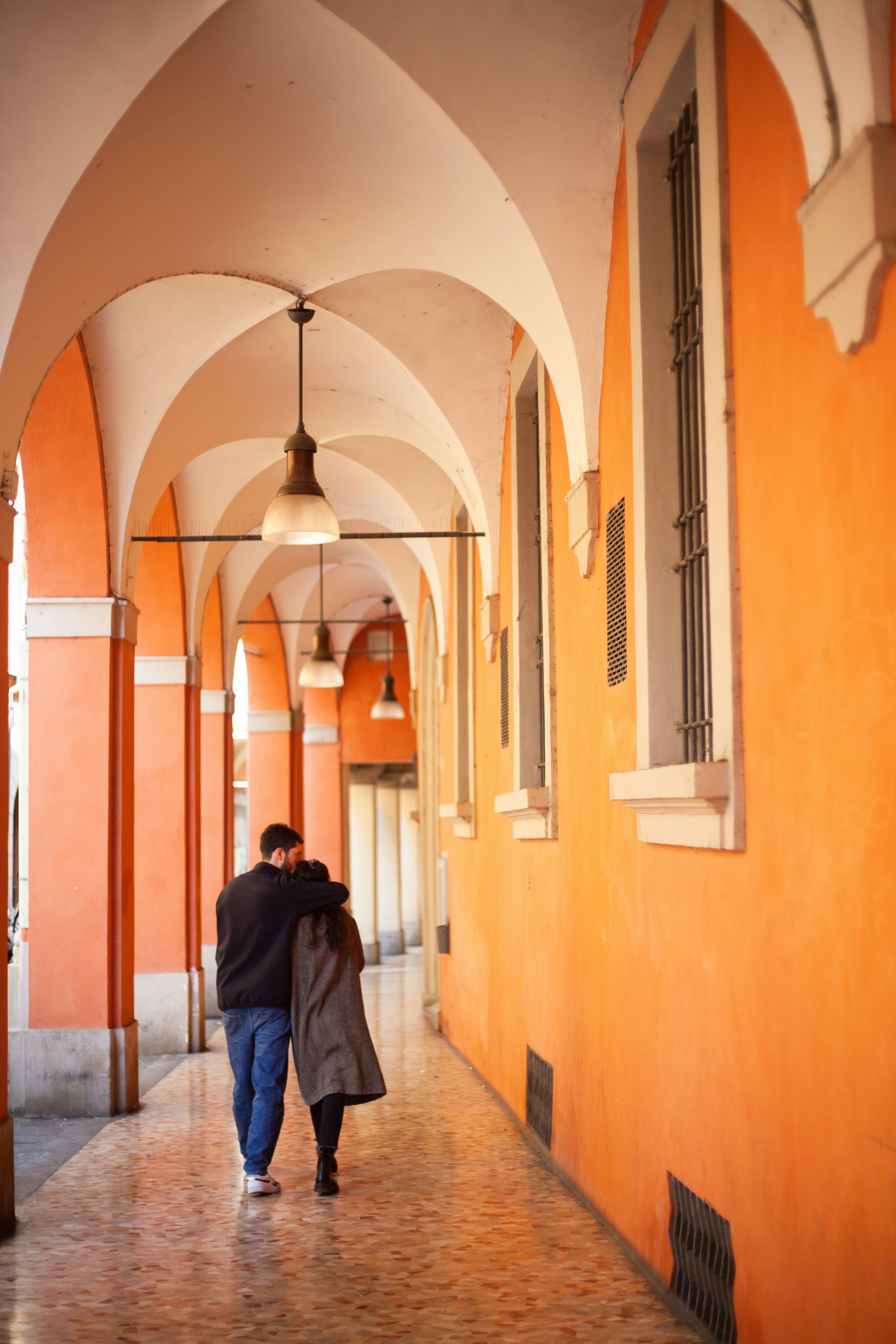 Couple walking closely together under archways in a vibrant orange corridor, evoking a sense of intimacy and connection.