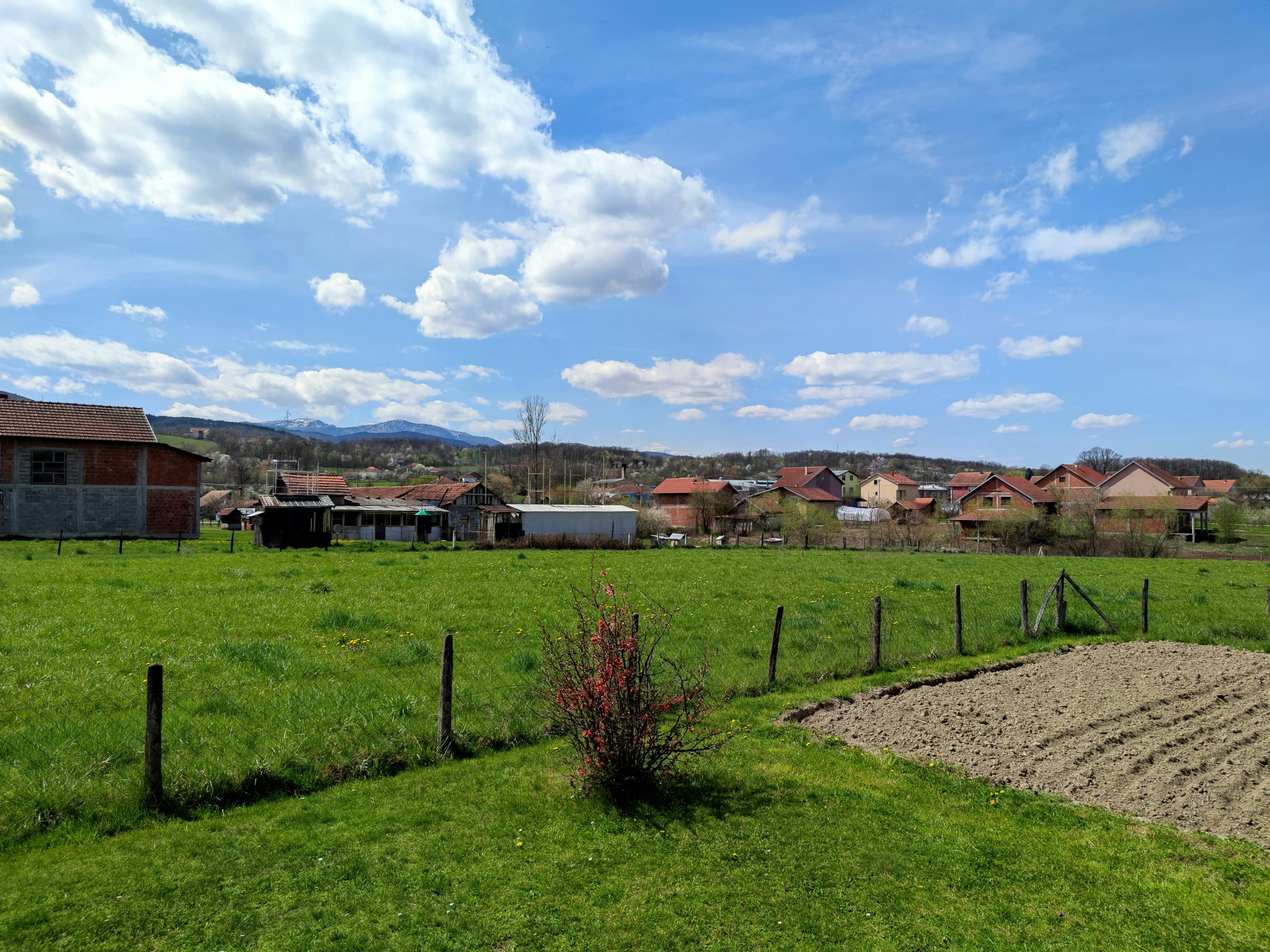Rural landscape photograph with a foreground shrub and fence guiding the eye toward distant houses beneath a bright, cloud-dotted sky.