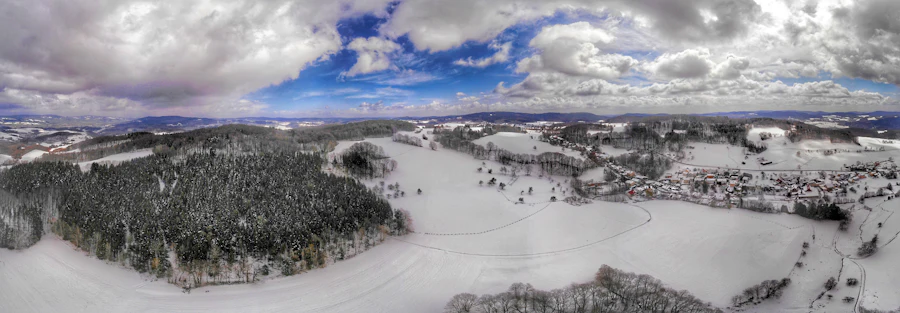 Wide aerial view of a snow-covered valley with dark pine forest, open fields, and a small village beneath dramatic clouds. Blue sky breaks through near the horizon.
