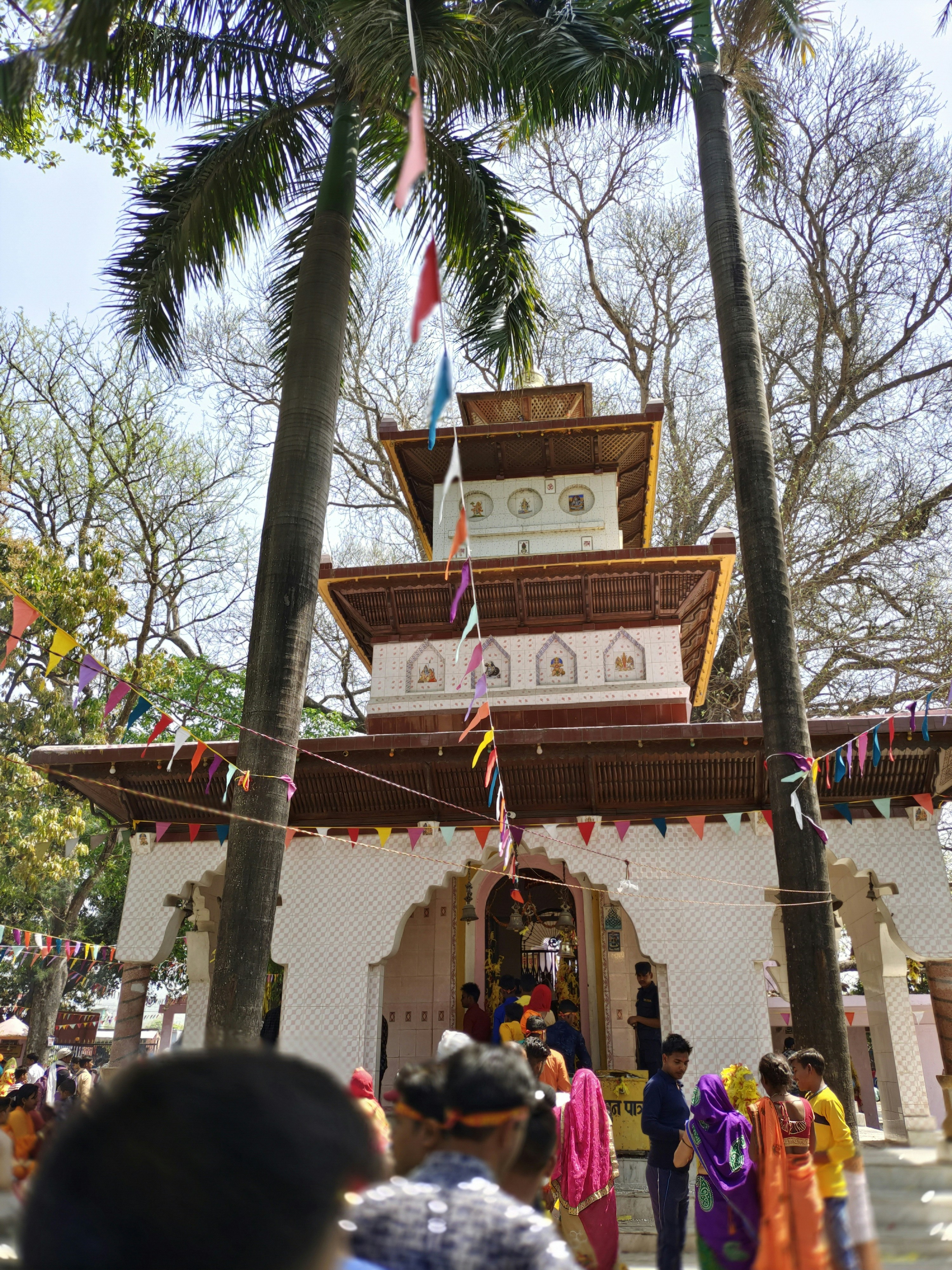 Colorful temple tower framed by palm trees and fluttering prayer flags, with a crowd gathered at the entrance during a festival.