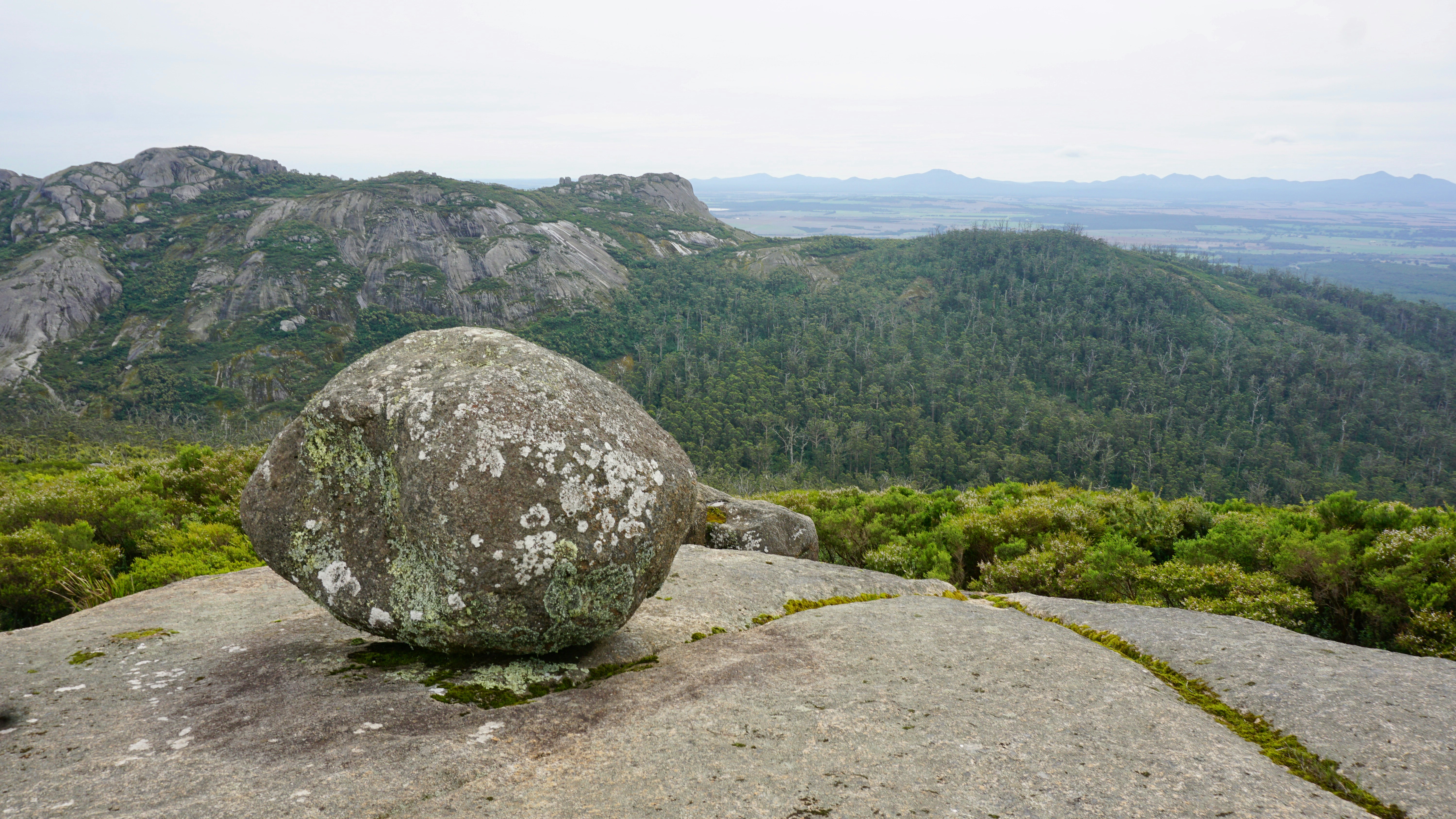 Lichen-covered boulder perched on a rocky outcrop overlooking a vast, green landscape with distant mountains.