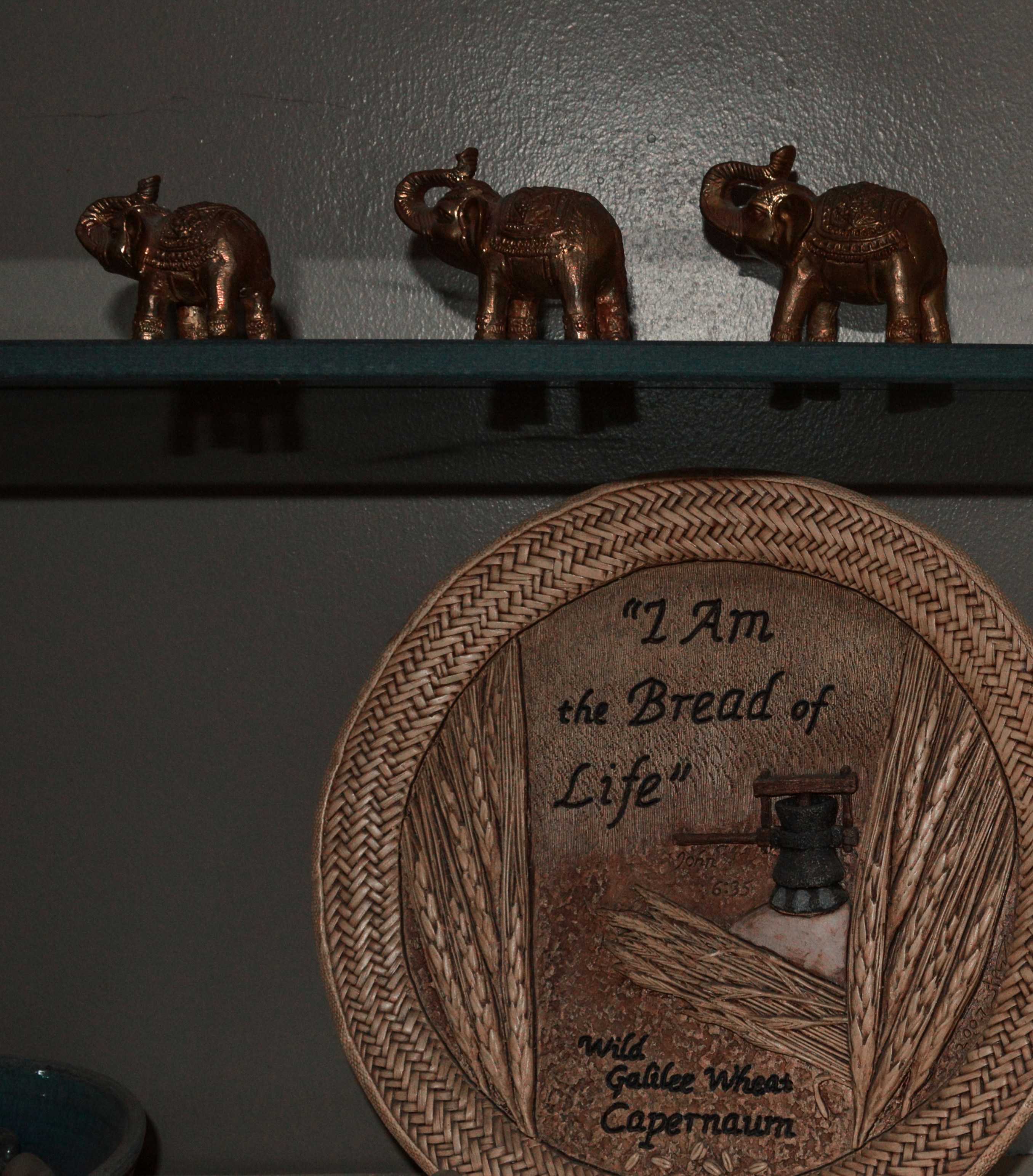 Decorative plate with the phrase 'I Am the Bread of Life' surrounded by intricate designs, accompanied by small elephant figurines on a shelf.