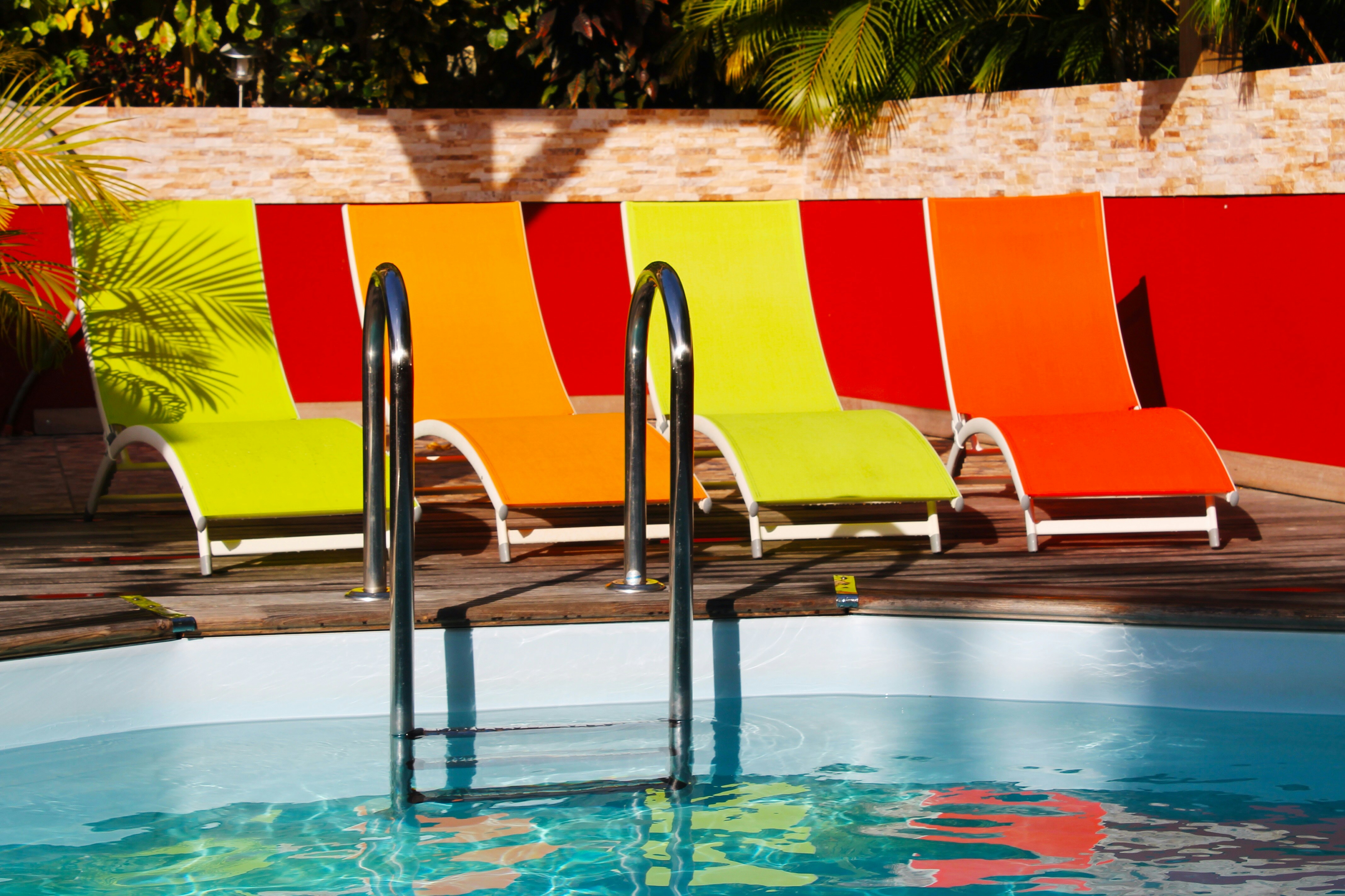 Colorful lounge chairs line the edge of a sparkling pool, surrounded by lush greenery and a vibrant red wall. The scene invites relaxation and enjoyment.