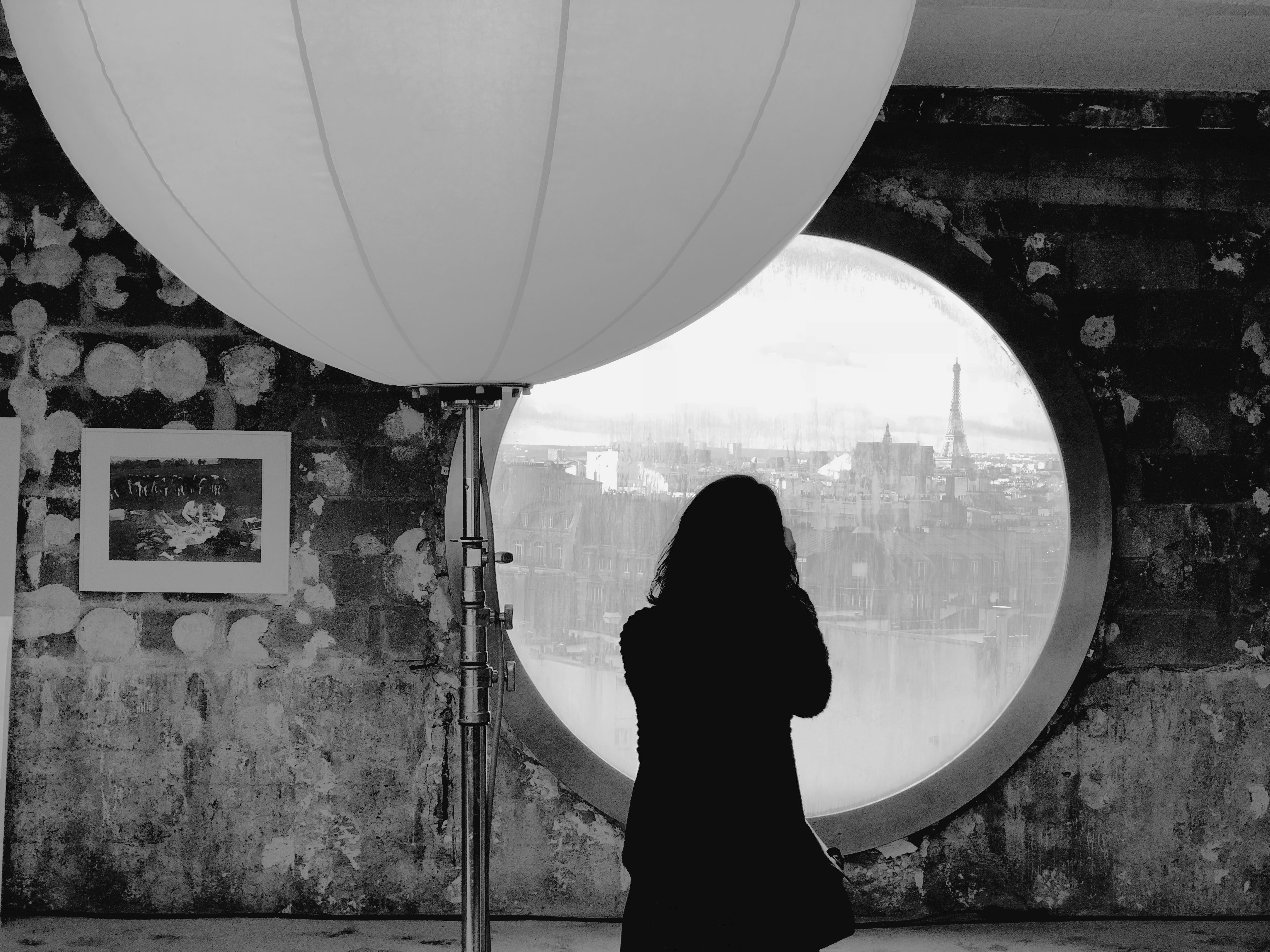 Silhouette of a person framed by a large circular window, overlooking a cityscape with the Eiffel Tower in the distance. A large balloon light source adds an artistic touch.