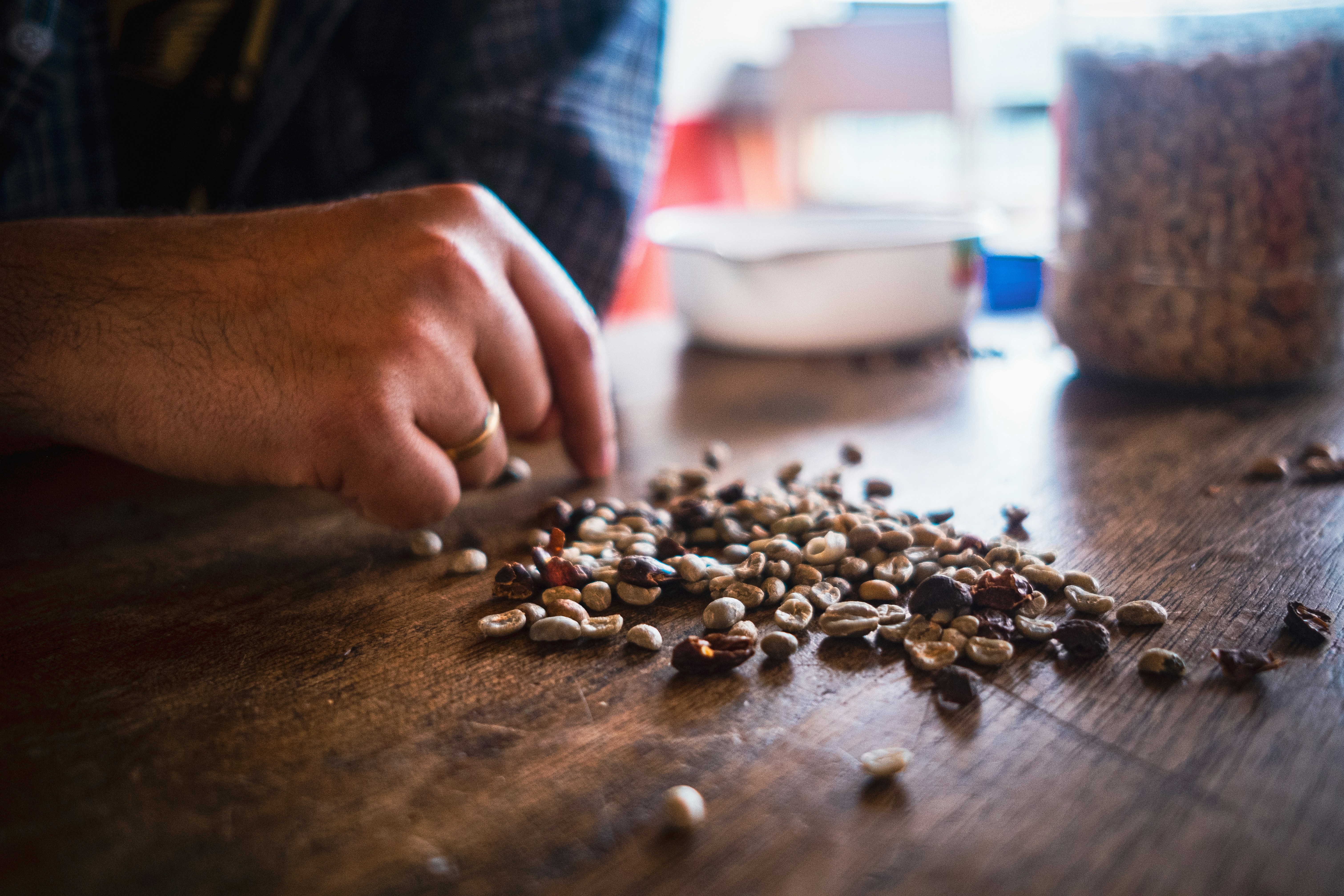 Hand arranging a mix of seeds on a wooden table with a jar and bowl in the background.