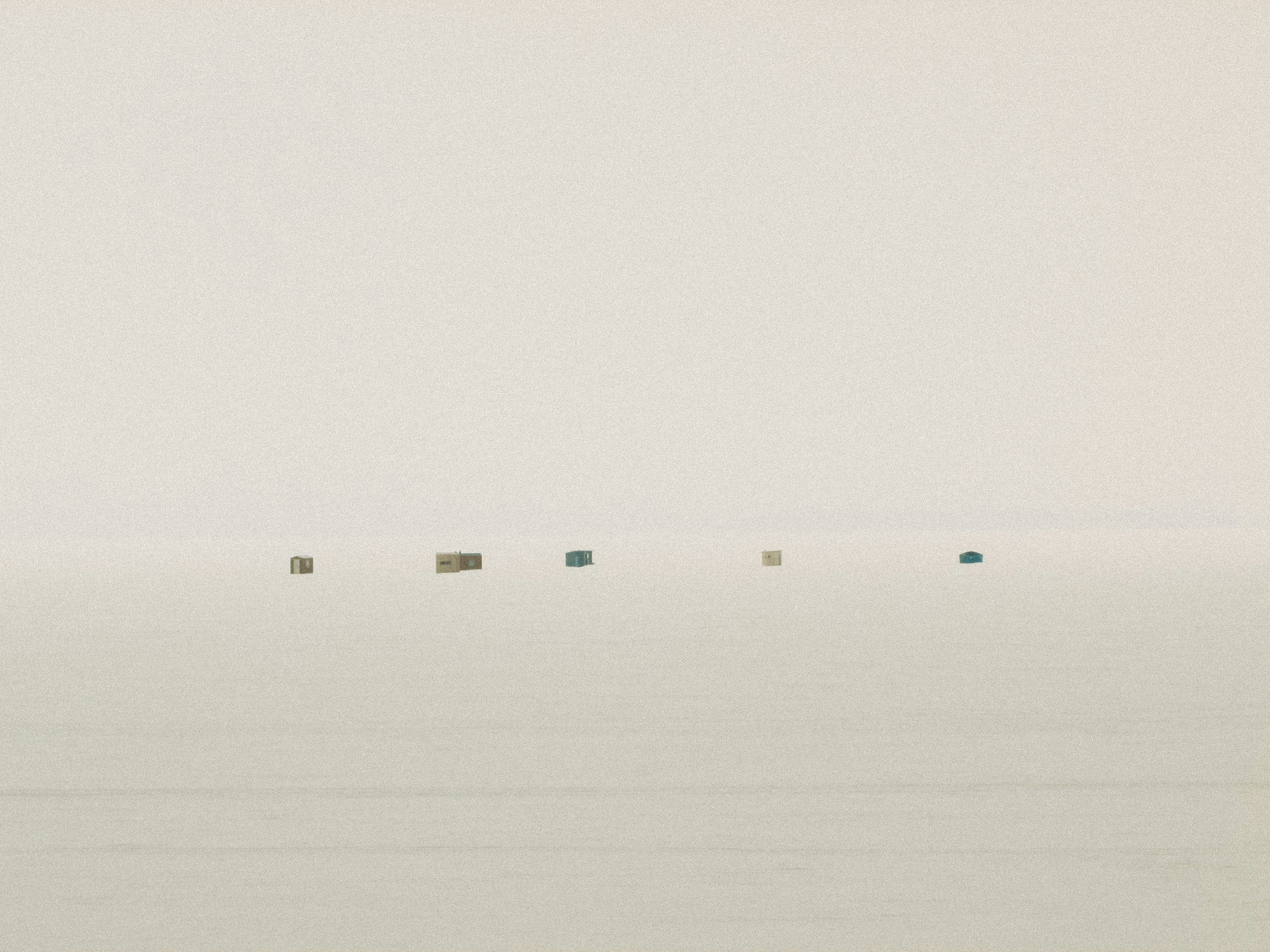 Small ice huts dot a vast, foggy horizon, blending into the muted landscape.