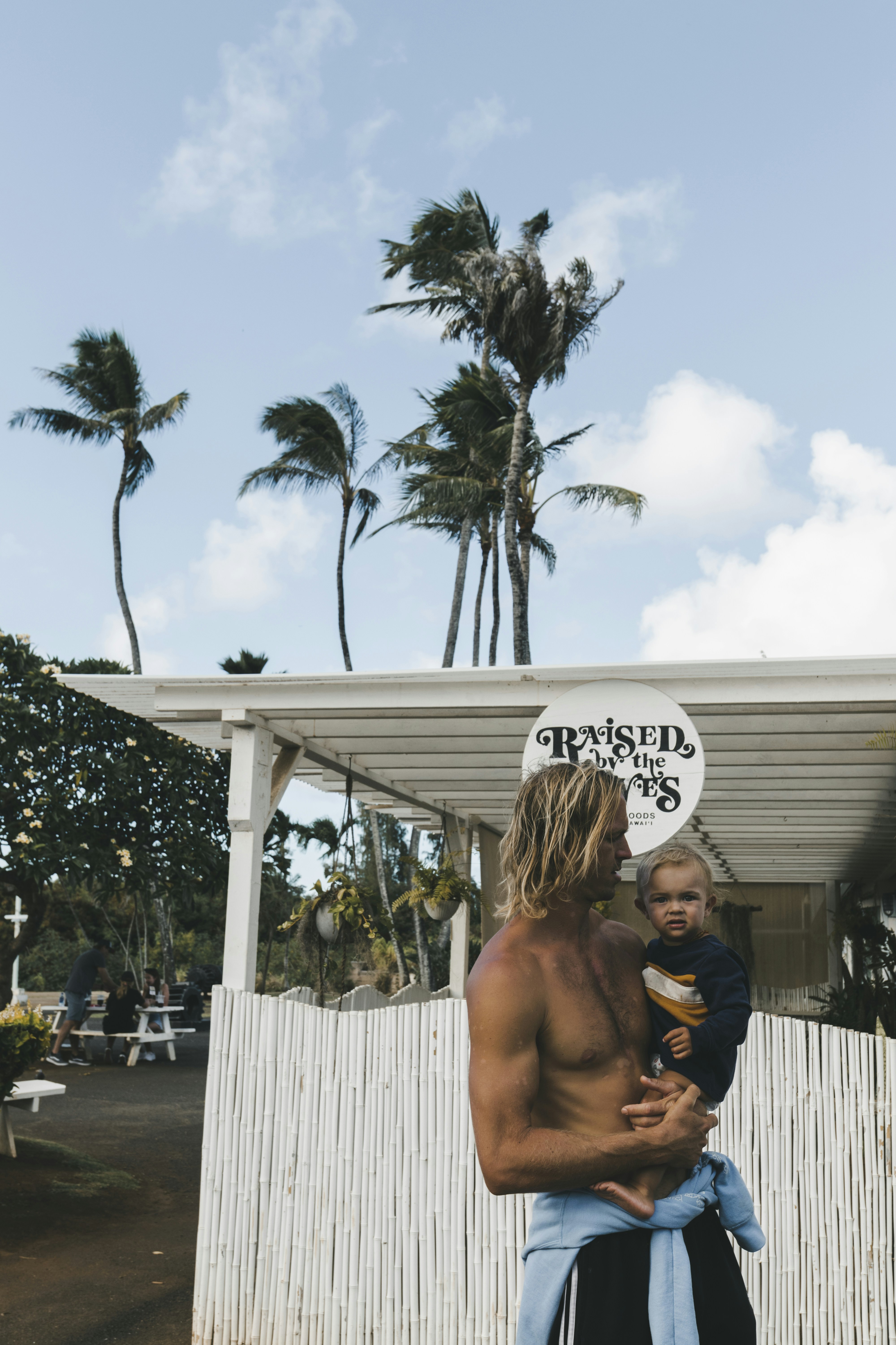 A man holds a child against a backdrop of swaying palm trees and a whimsical sign, capturing a serene moment in a tropical setting.