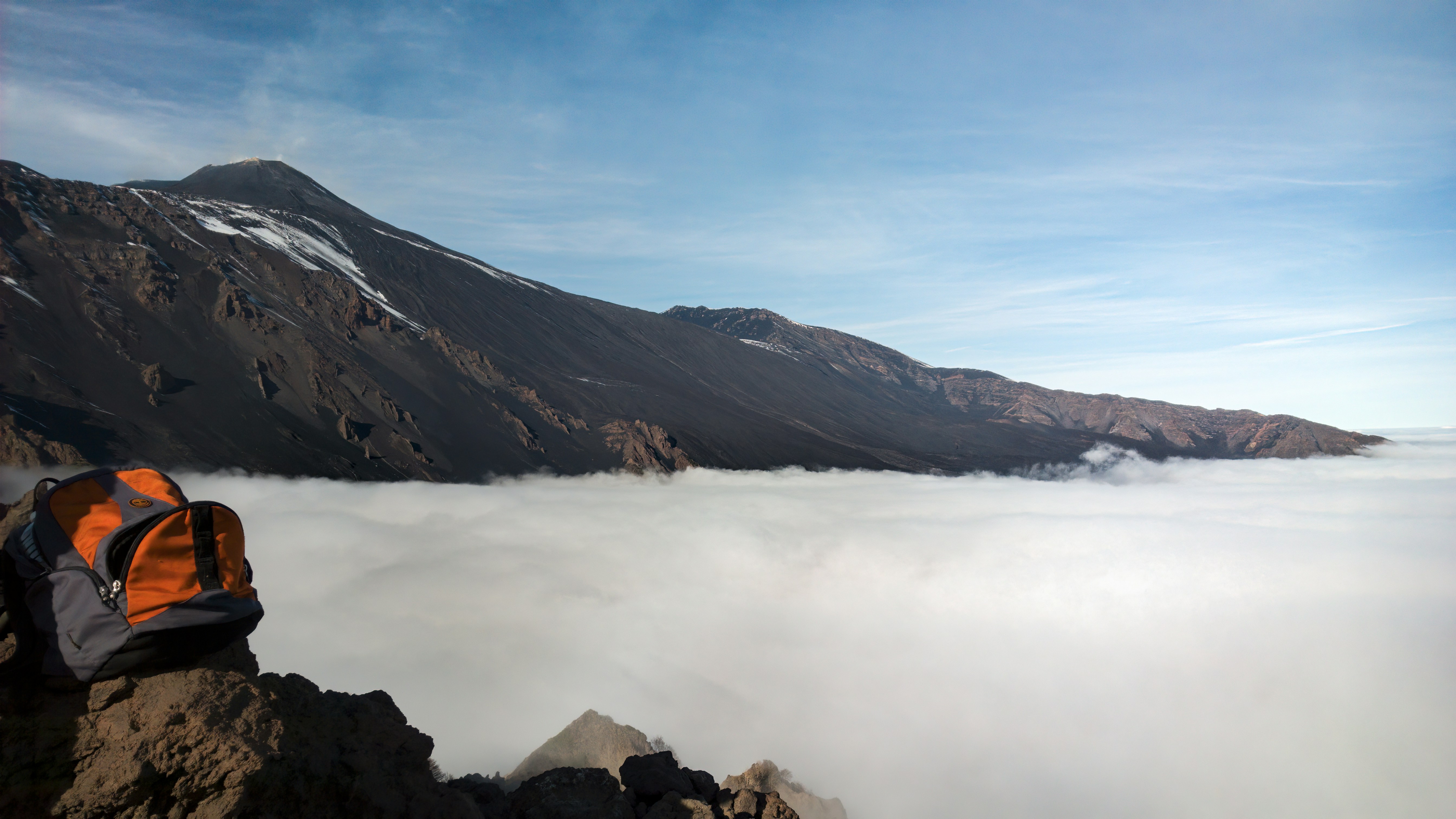 Hiker's backpack resting on rocky terrain, overlooking a sea of clouds with a volcanic mountain in the background. 