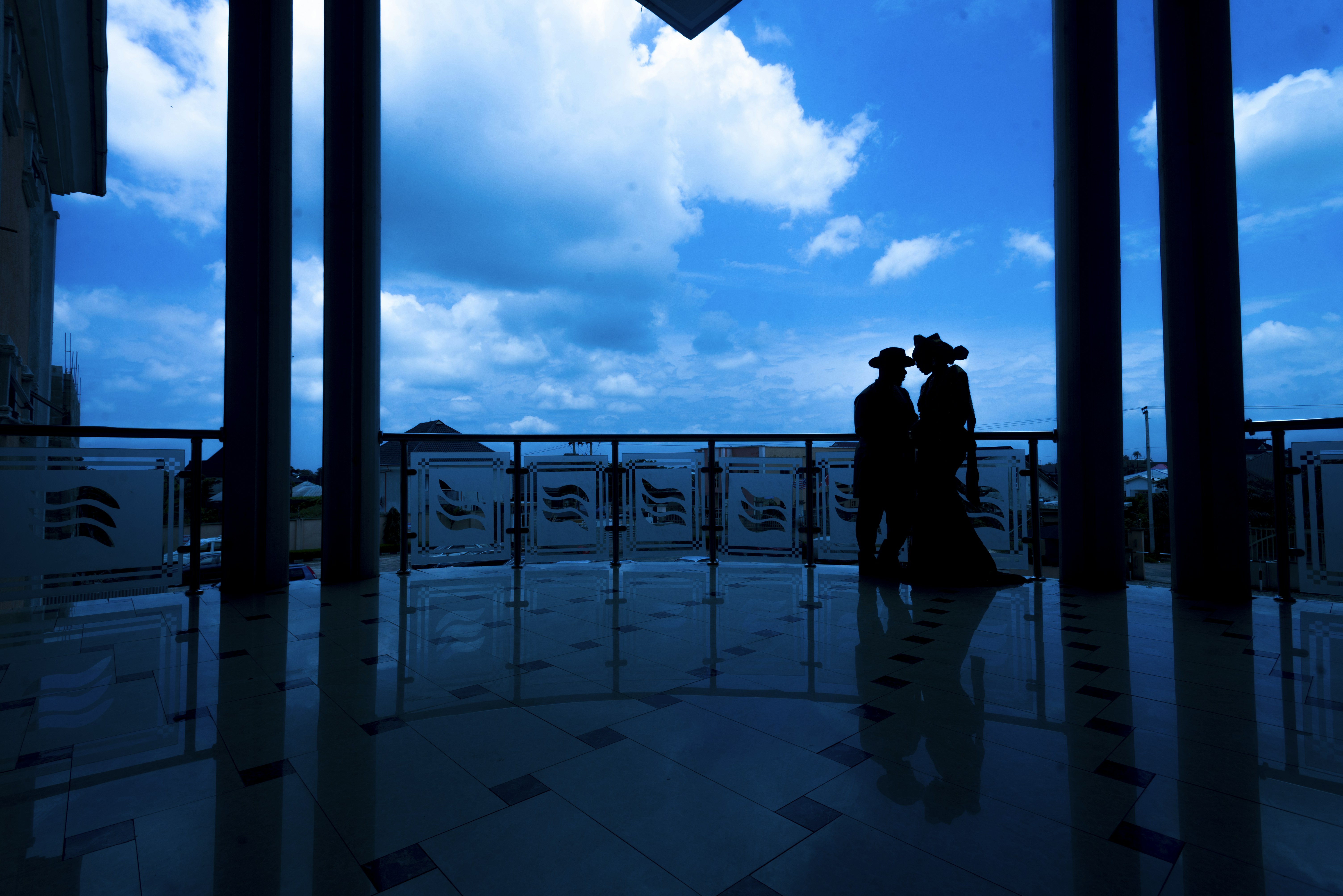 Silhouetted figures stand under a vast blue sky on a balcony with tall columns.