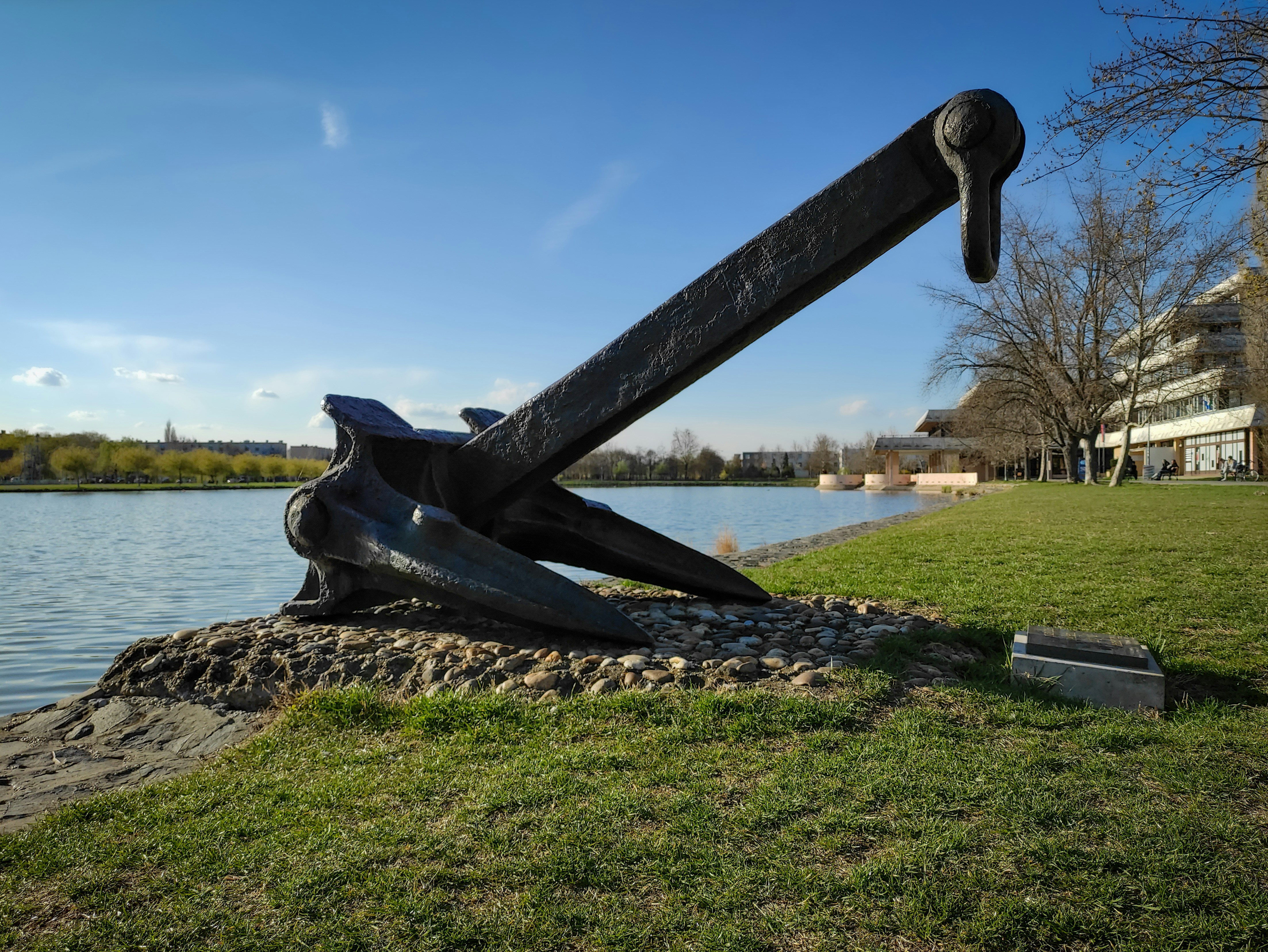 Massive black anchor resting on grassy bank beside a tranquil river under clear blue sky.