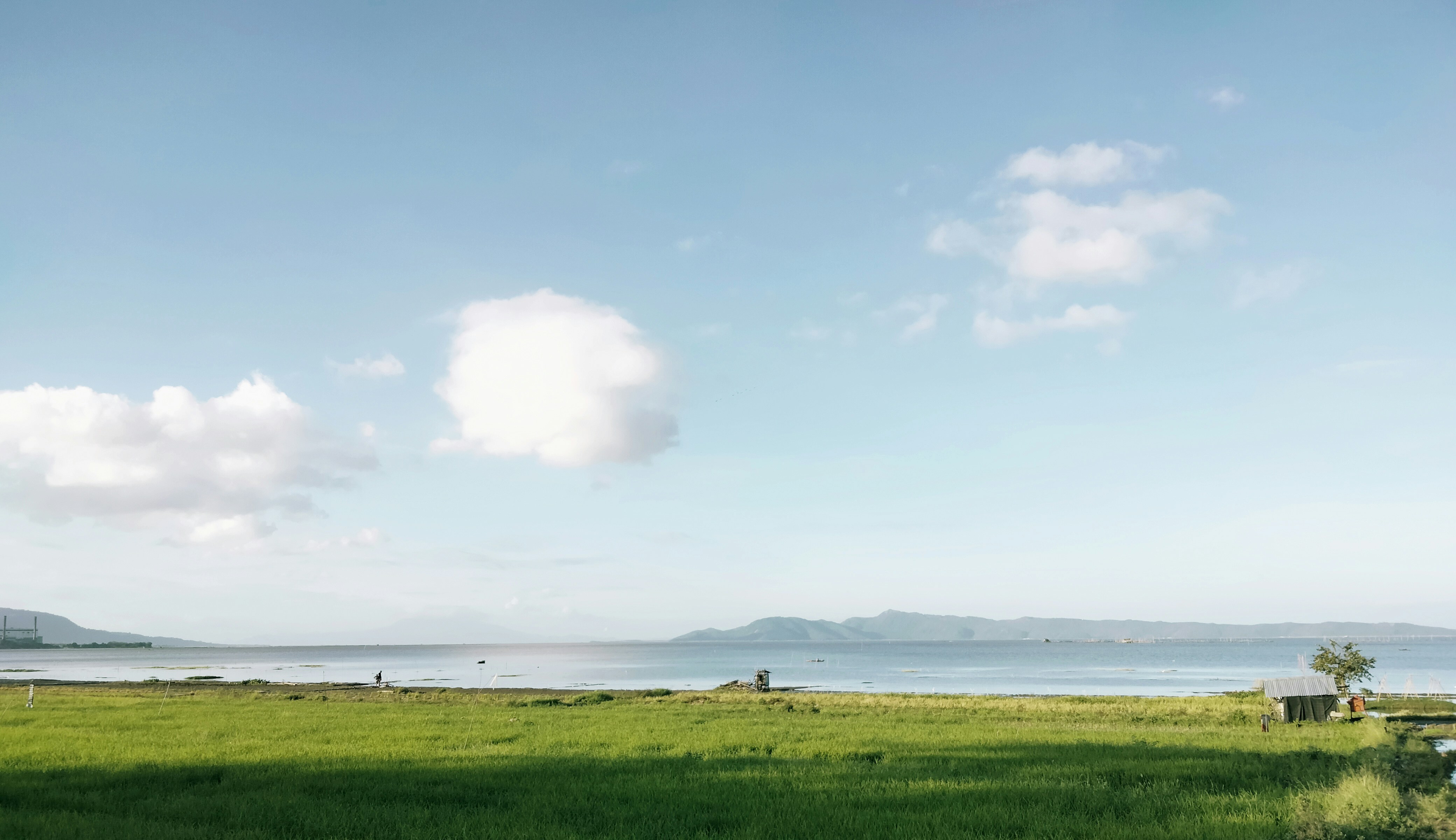 Expansive green field leading to a tranquil lake under a bright blue sky with scattered clouds.