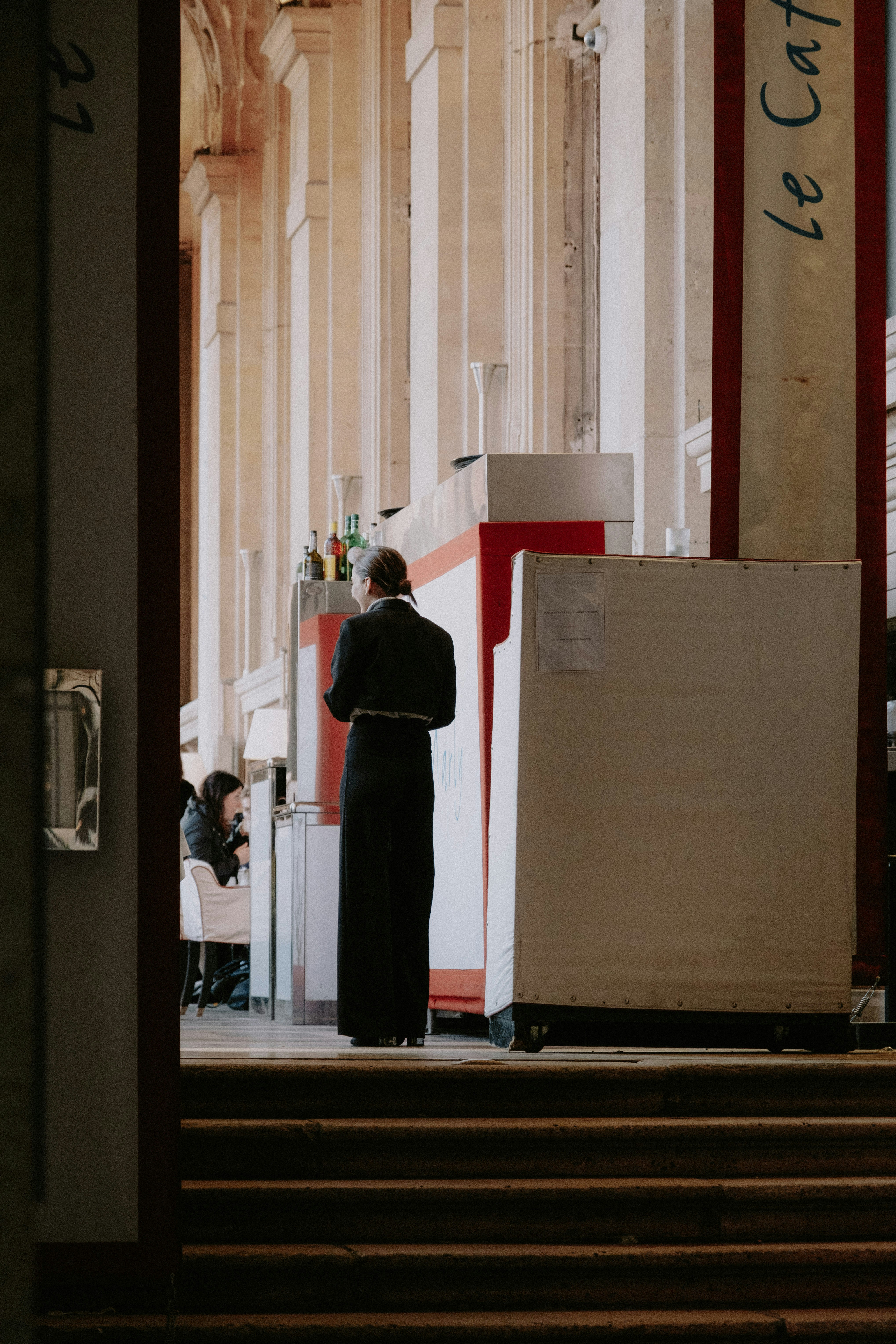 Silhouette Among Columns: A solitary figure near a red kiosk in a grand arcade