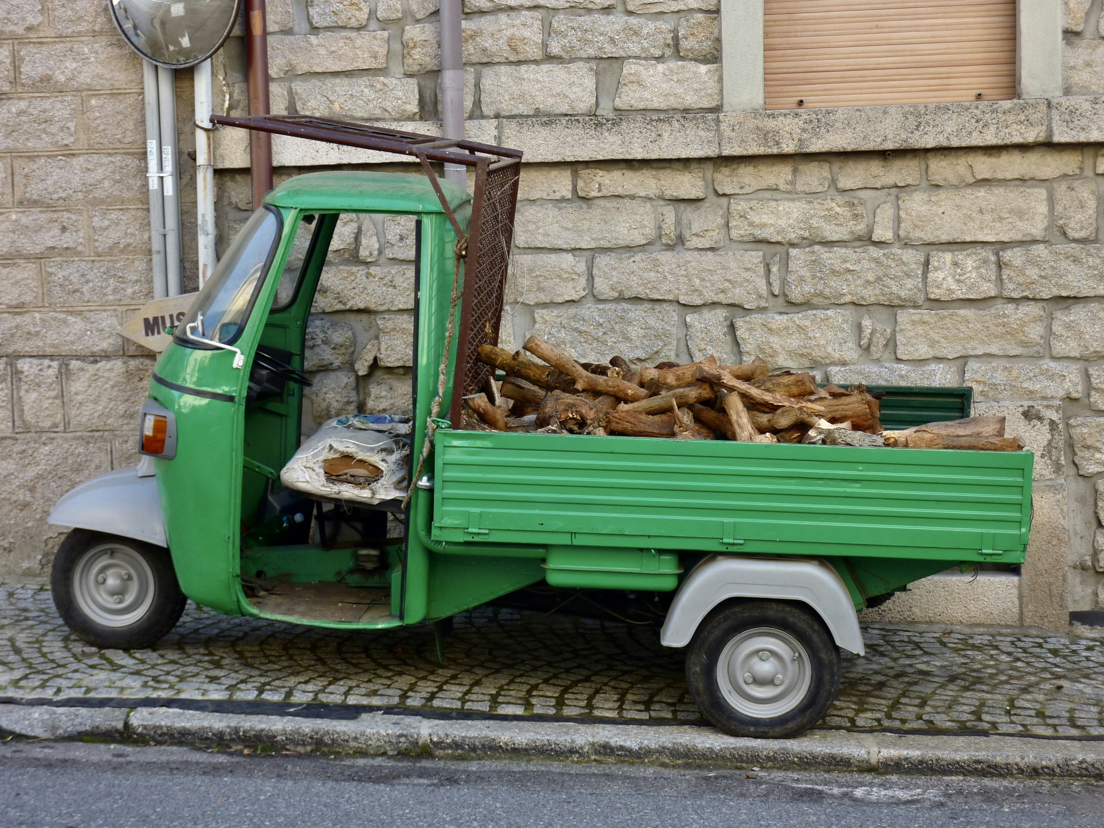 A green three-wheeled vehicle loaded with logs parked beside a stone wall, showcasing a blend of utility and charm.
