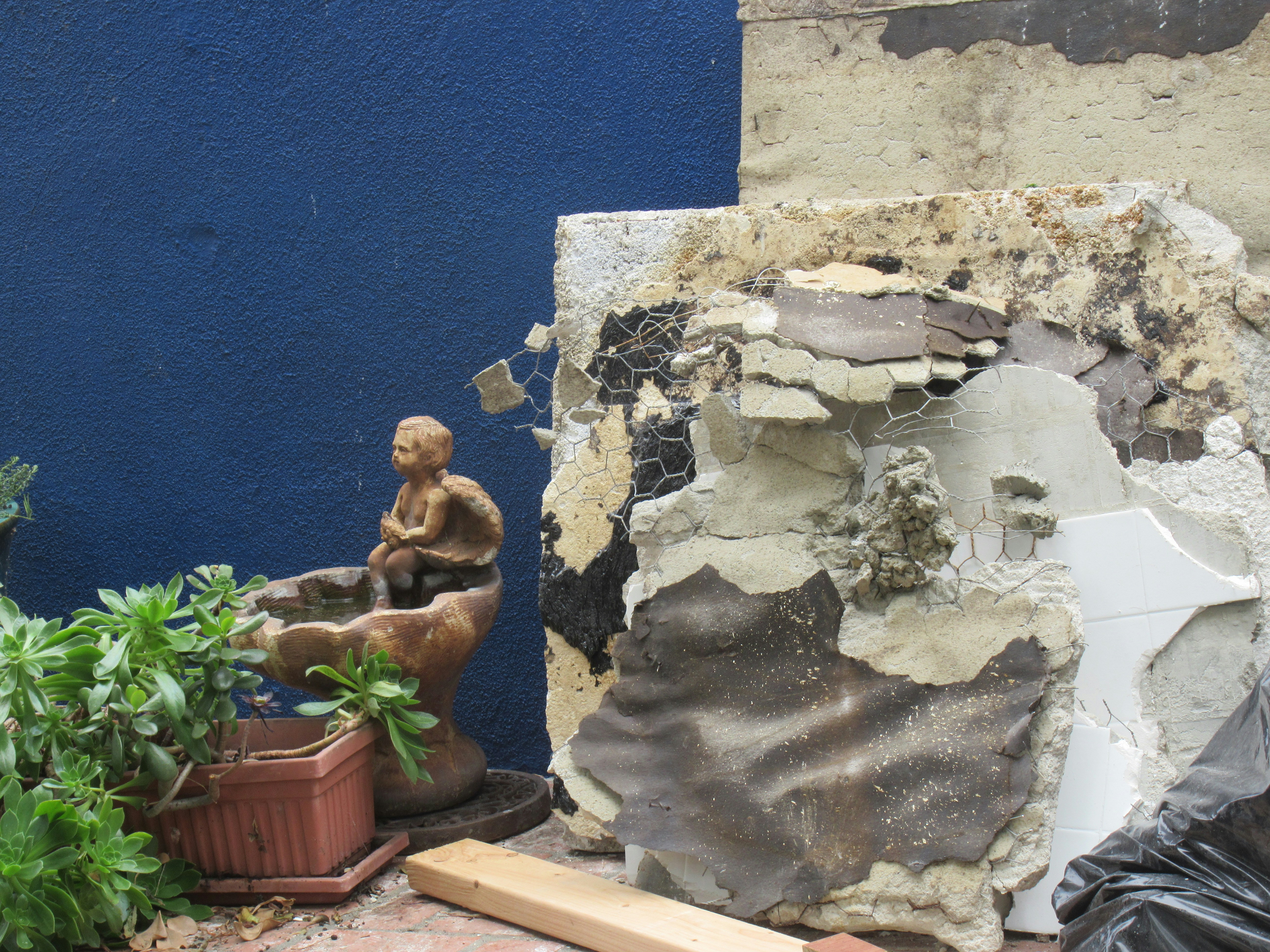 Cherub on a birdbath next to plaster demolition, considering the state of the world | a statue of a man sitting on a rock next to a potted plant