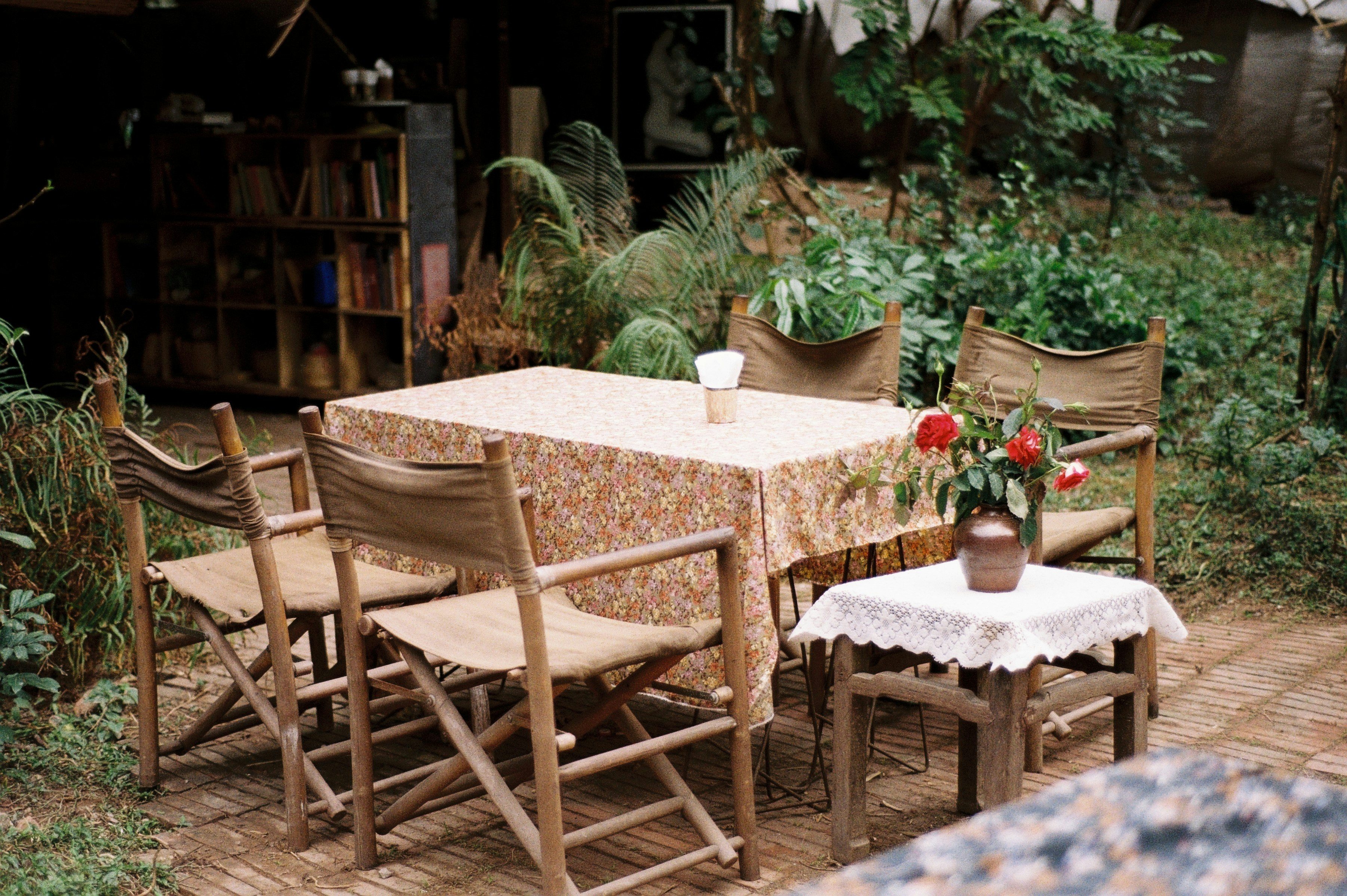 Wooden chairs around a table with a floral tablecloth in a garden, surrounded by lush greenery and a small side table with a potted plant.