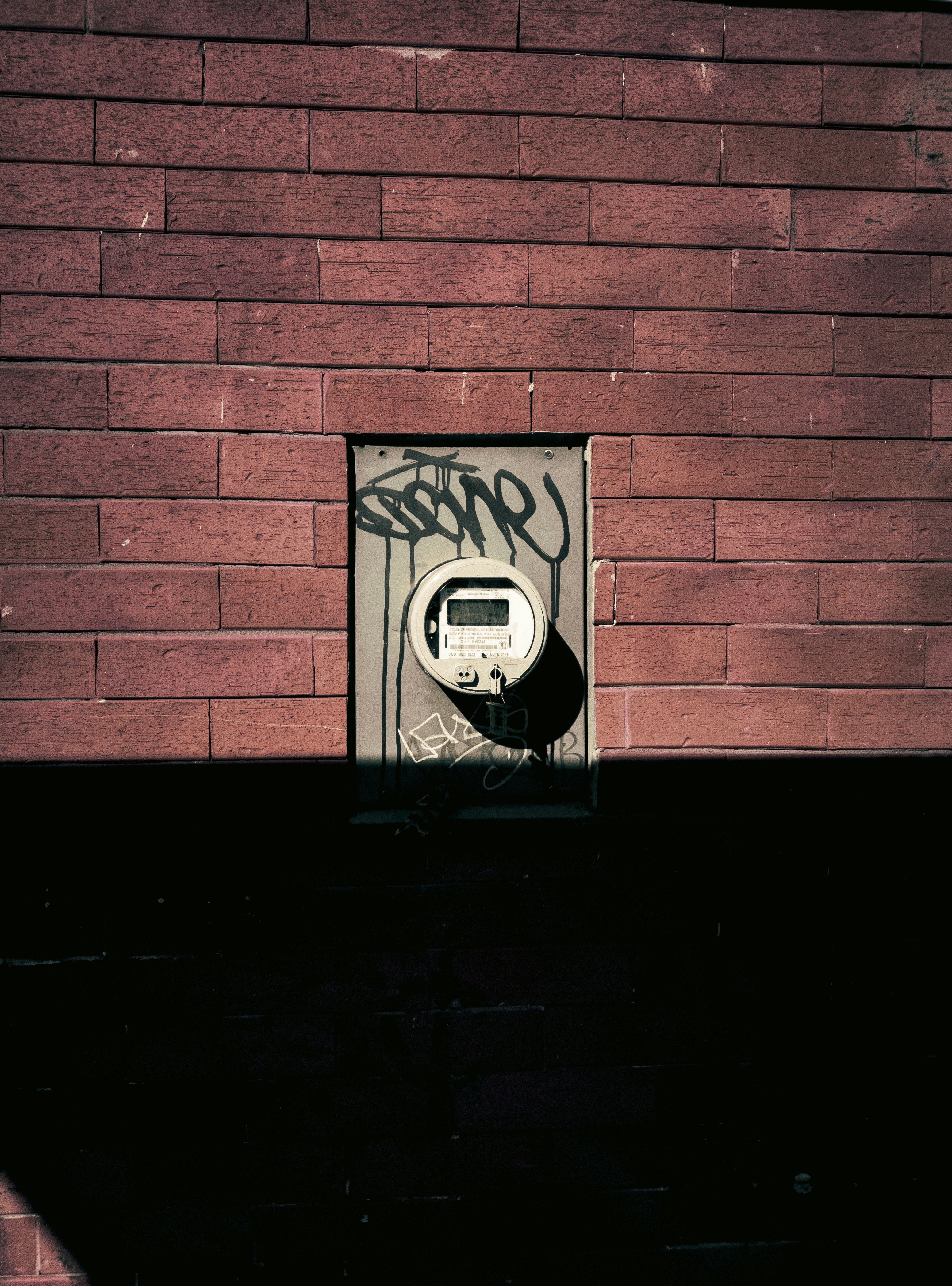 A street photograph of a graffiti-tagged utility meter set into a small, framed panel on a red brick wall. Harsh light creates a stark contrast with a deep shadowed band below, emphasizing texture.