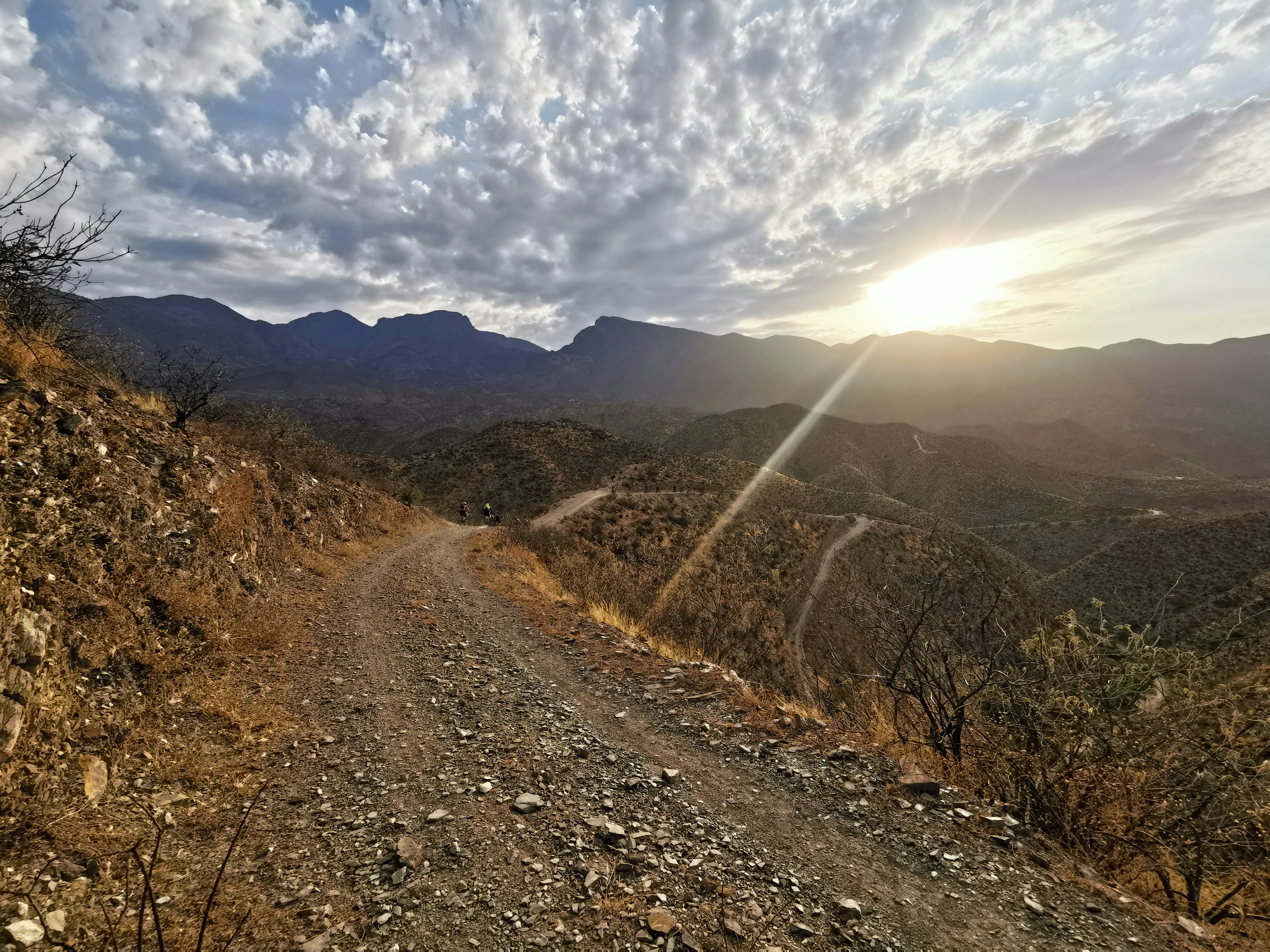 Rocky path winding through arid mountains under a dramatic, cloud-filled sky with the sun setting on the horizon.