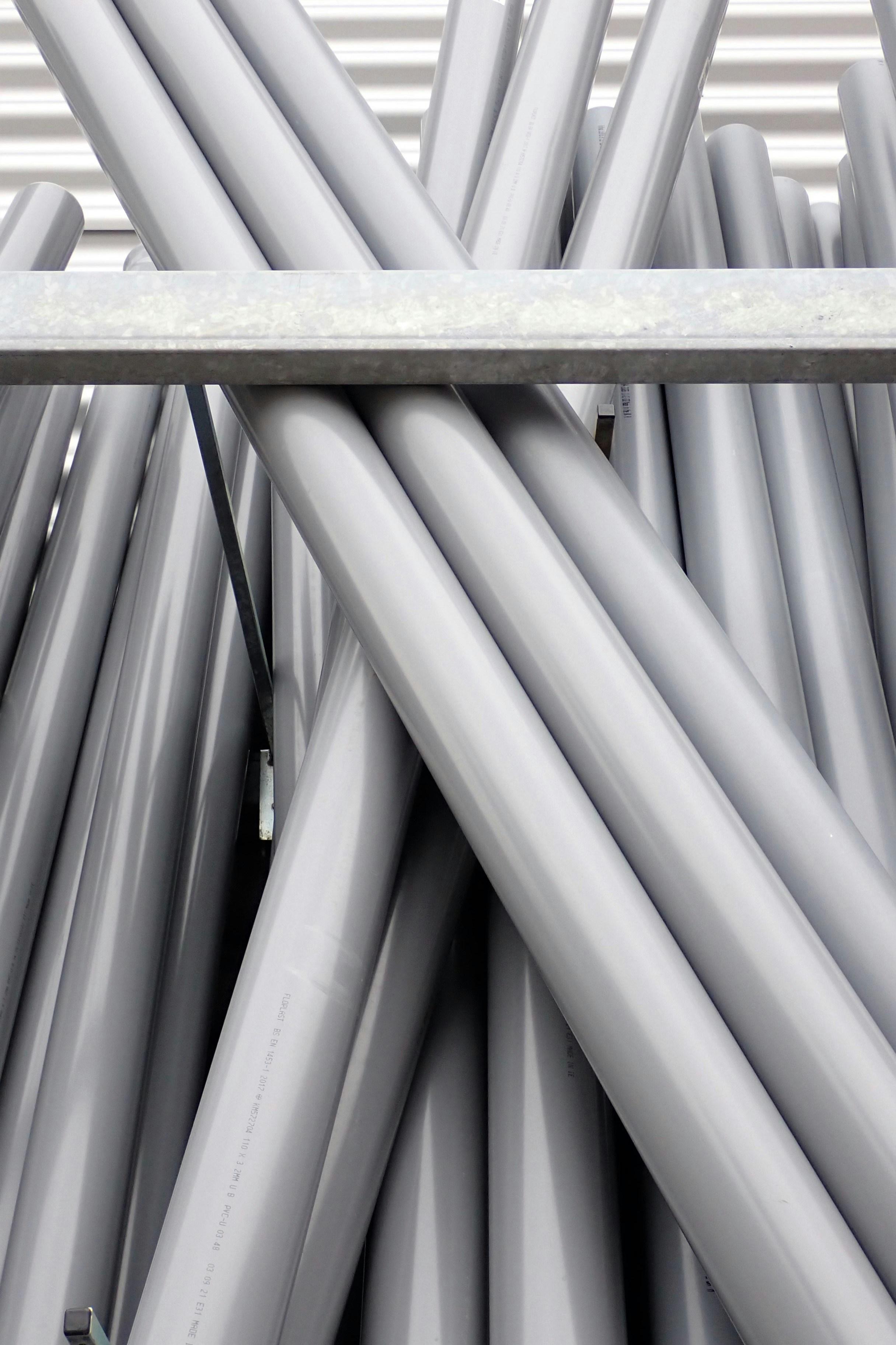 A chaotic arrangement of gray pipes stacked and intersecting, highlighting the complexity of industrial materials. The image captures the interplay of light and shadow on the smooth surfaces.