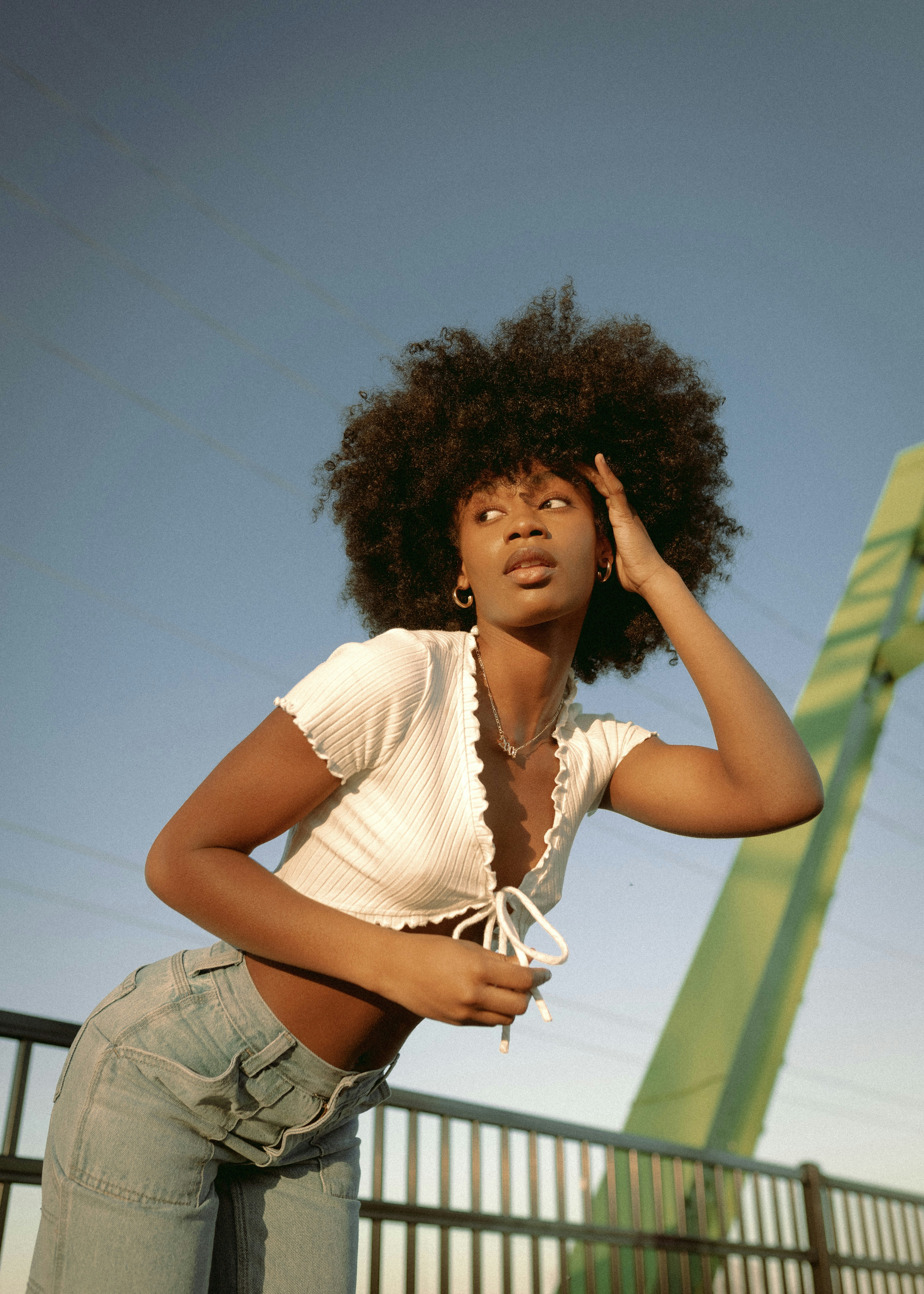 A young woman poses gracefully against a vibrant green bridge, her natural curls catching the light. The scene captures a blend of urban style and personal expression.