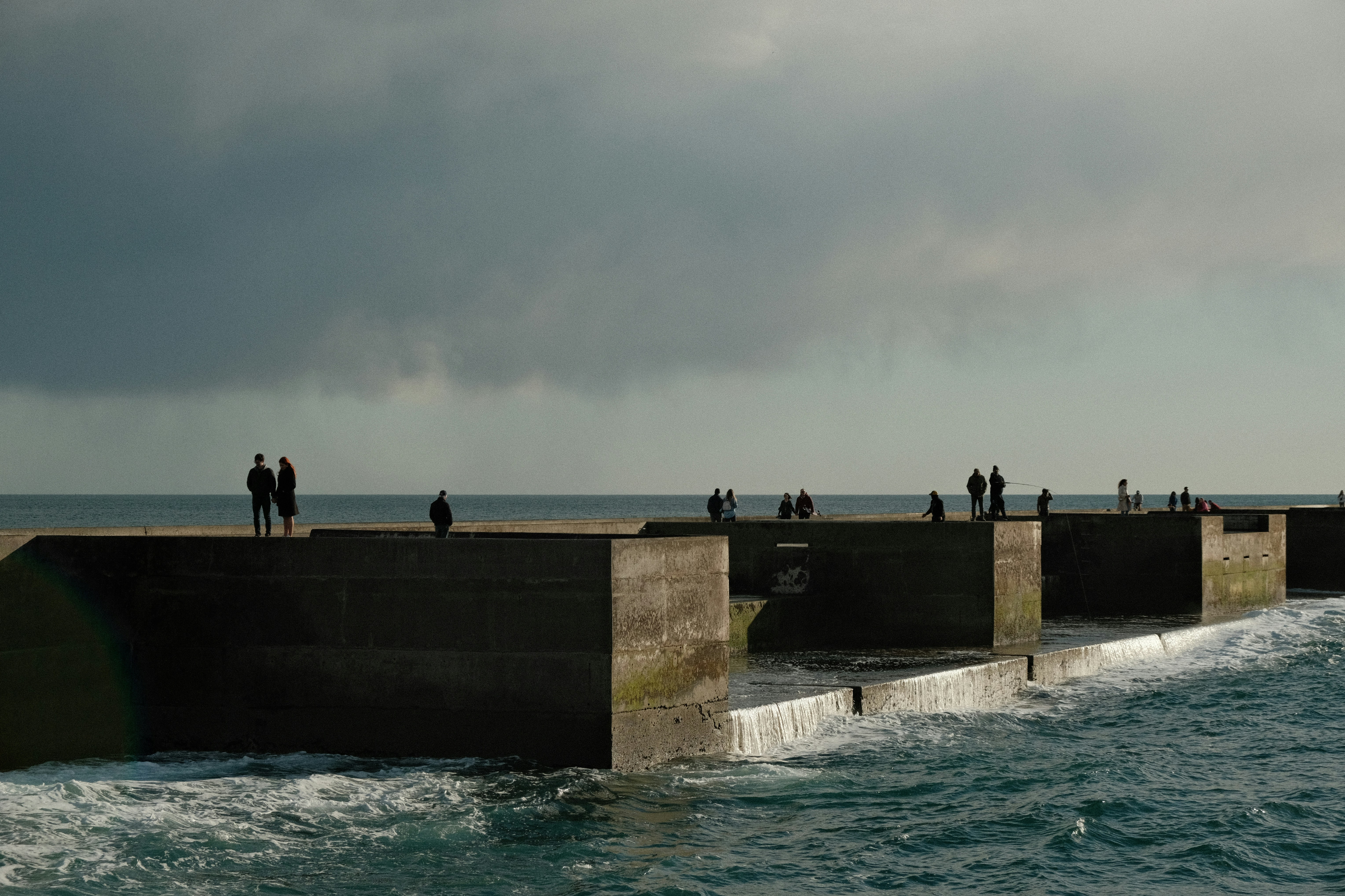 Concrete Promenade: Figures on a Pier Under a Moody Sea Sky