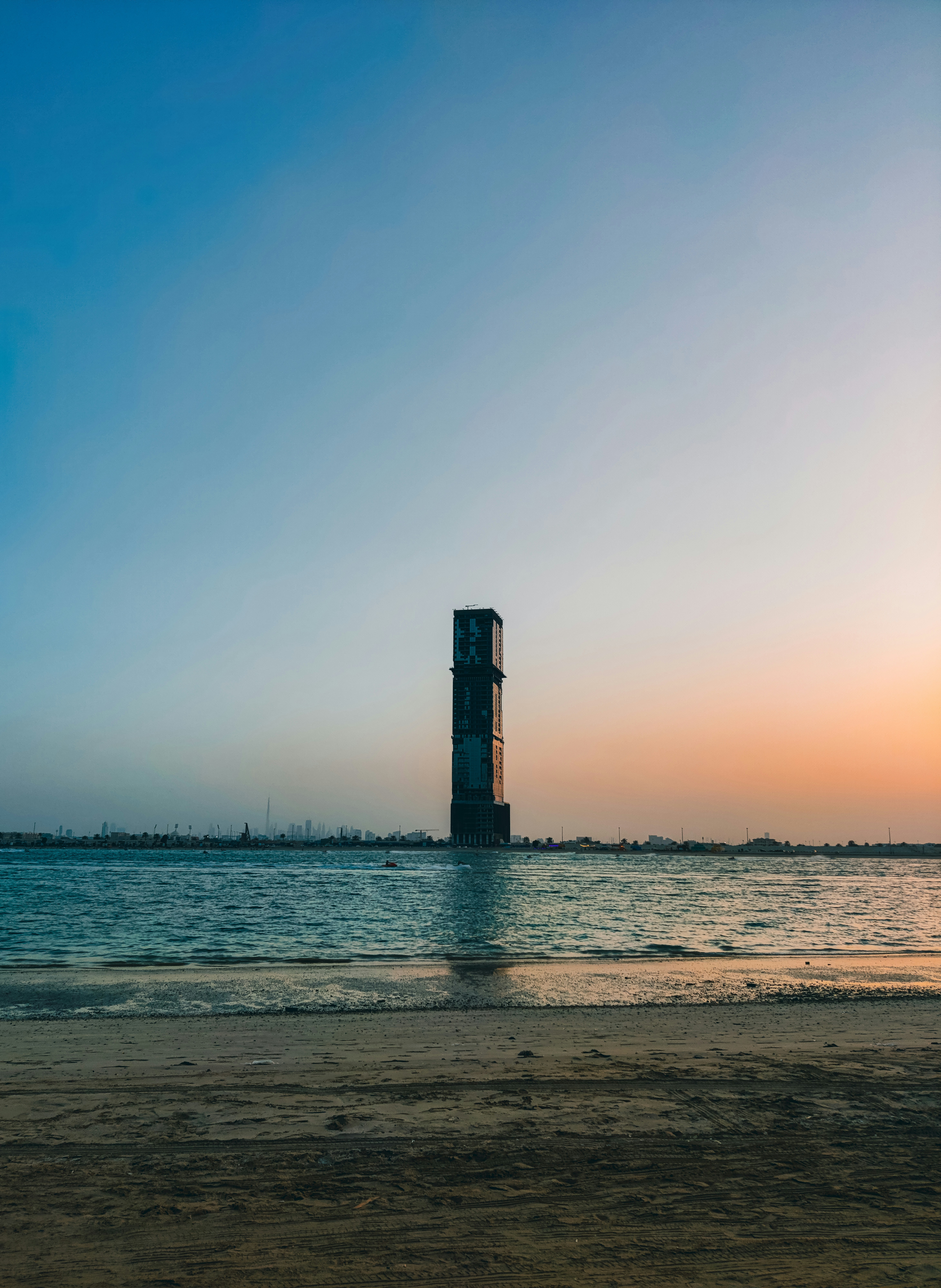 A towering structure rises from the water's edge against a gradient sky transitioning from blue to orange. The calm surface reflects the silhouette of the tower.