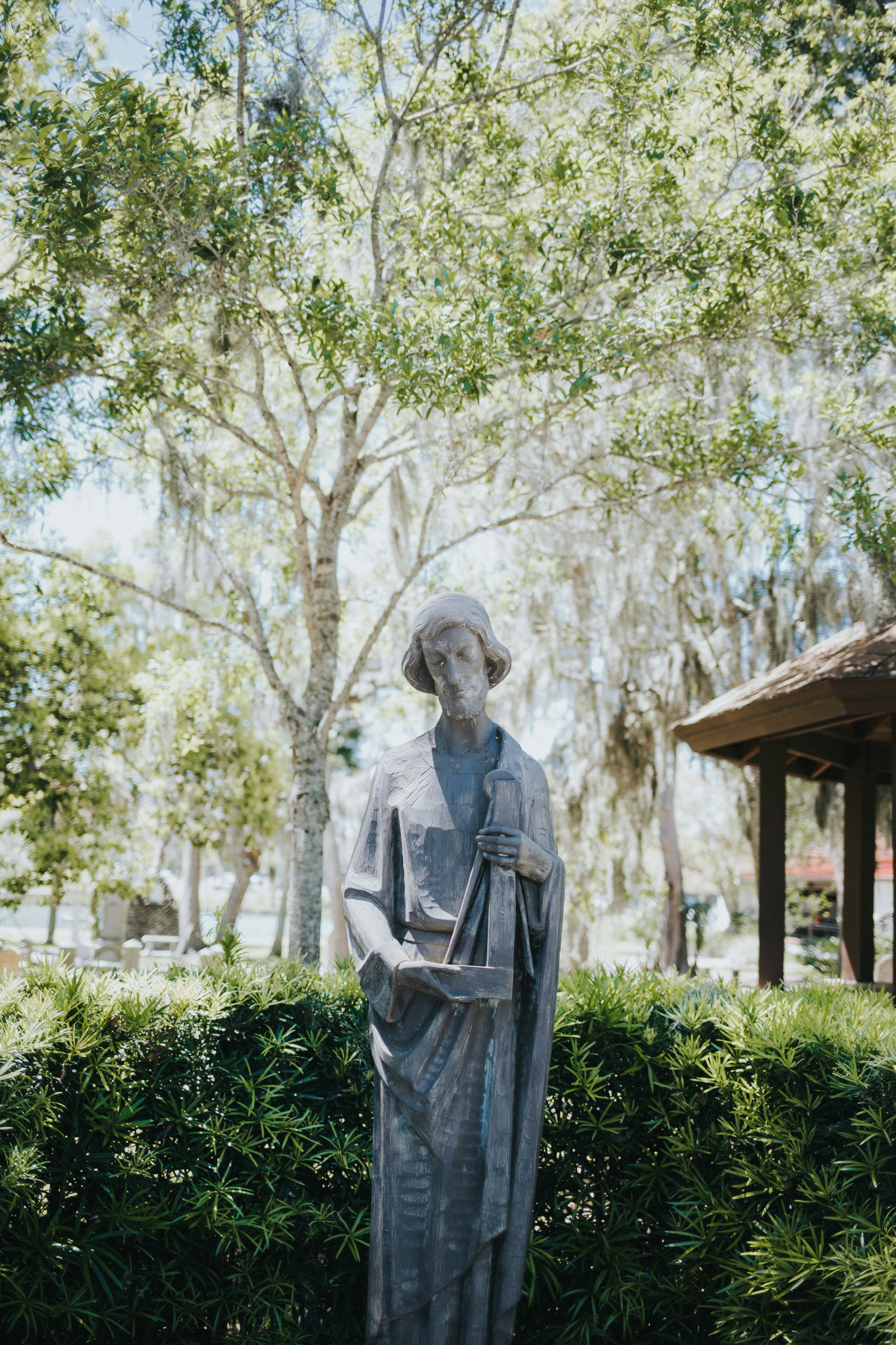 A statue of a figure holding a book and a tool, surrounded by lush greenery and trees, evoking a sense of wisdom and tranquility.
