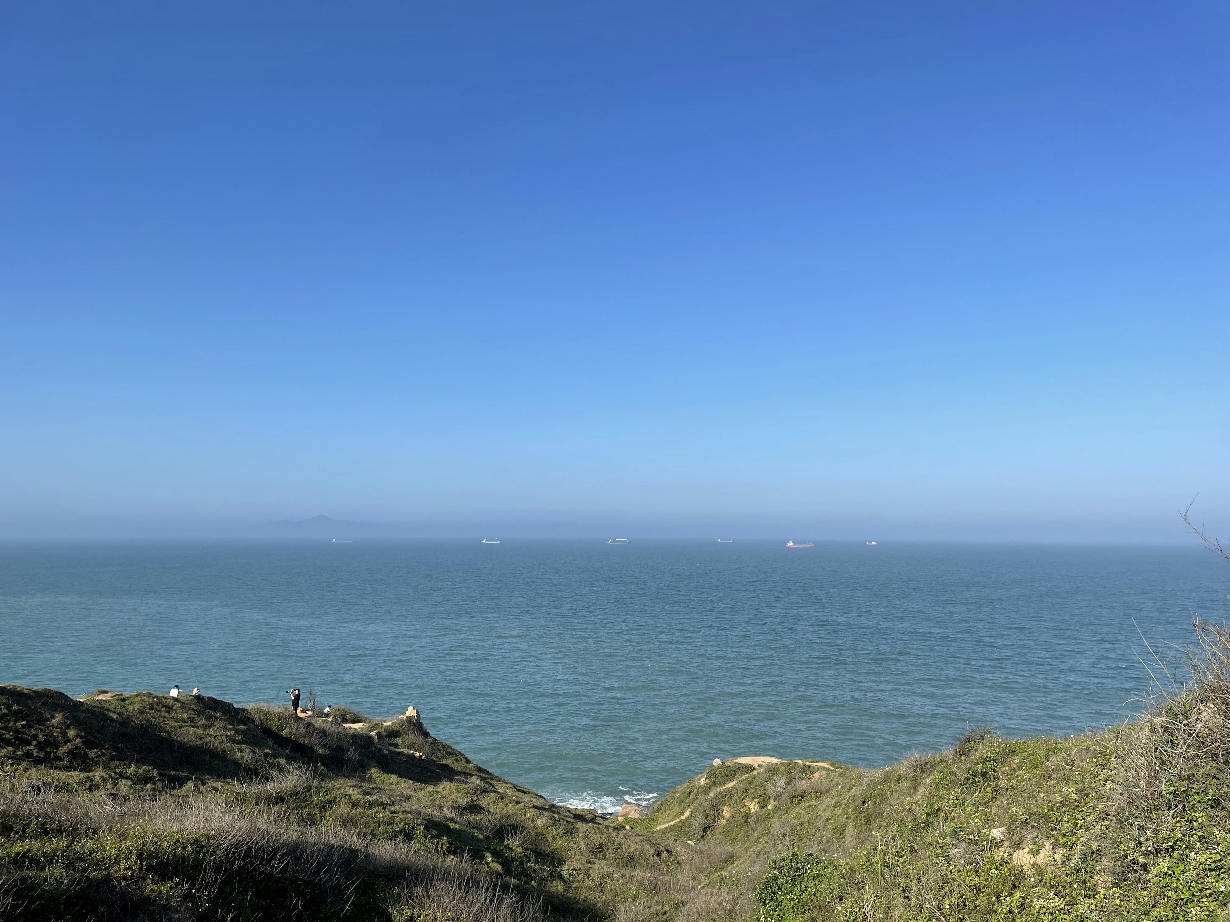 Expansive view of a tranquil ocean with distant ships under a clear blue sky. The lush green hillside frames the scene.