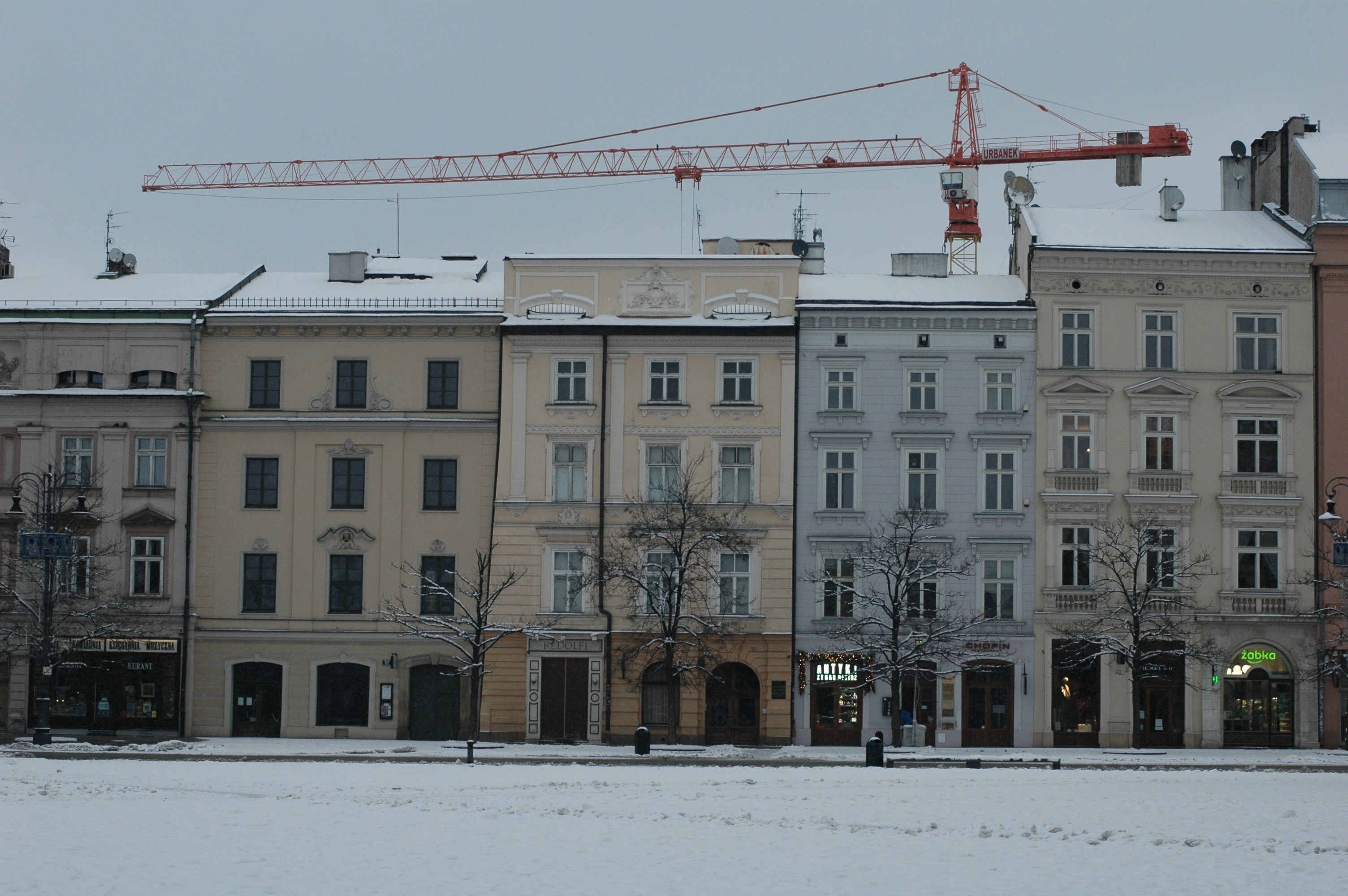 Row of historic buildings blanketed in snow, with a construction crane looming overhead in a gray winter sky.