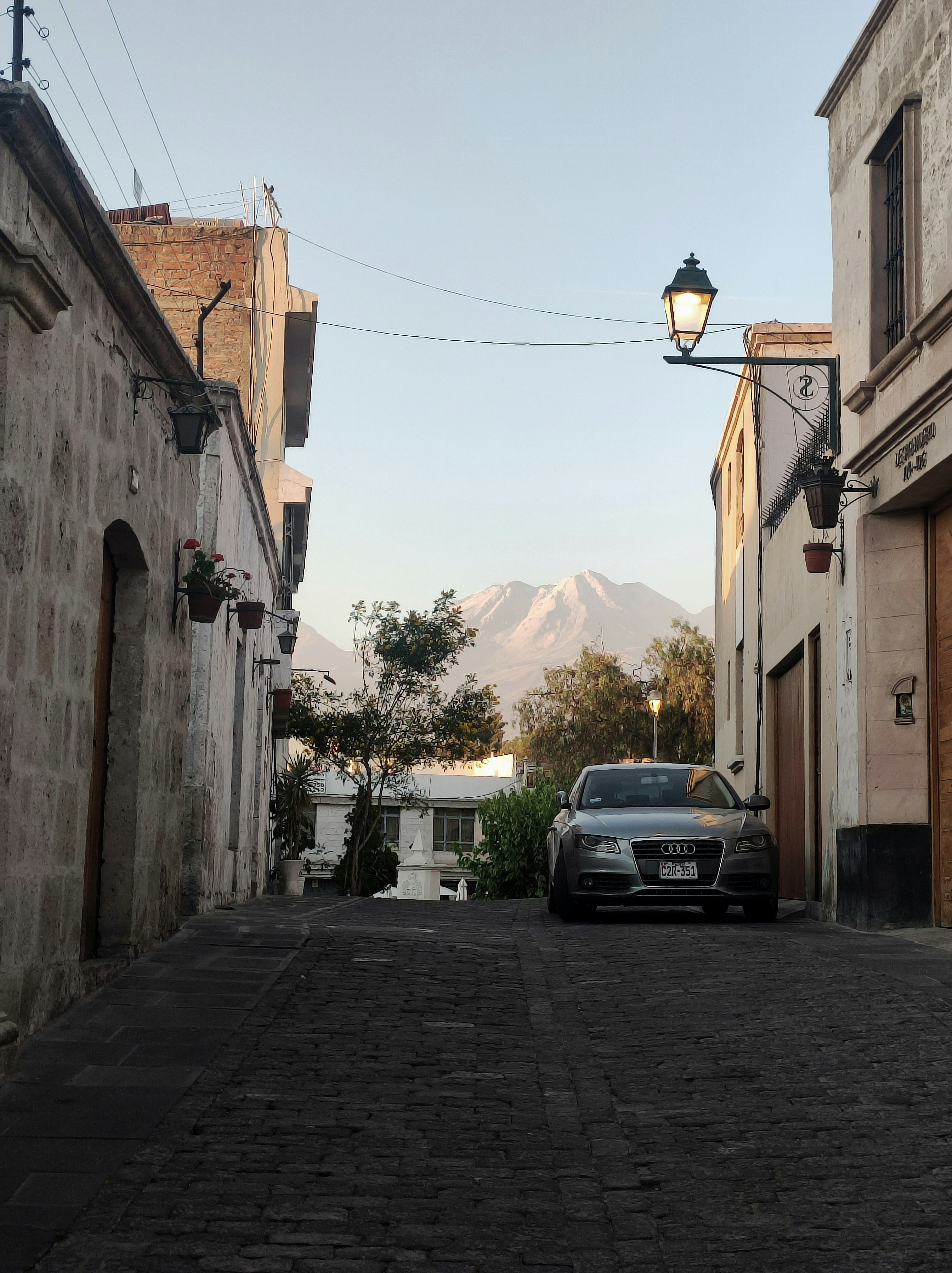 Narrow cobblestone street flanked by rustic buildings and a parked car, with a distant mountain peak under a clear sky.