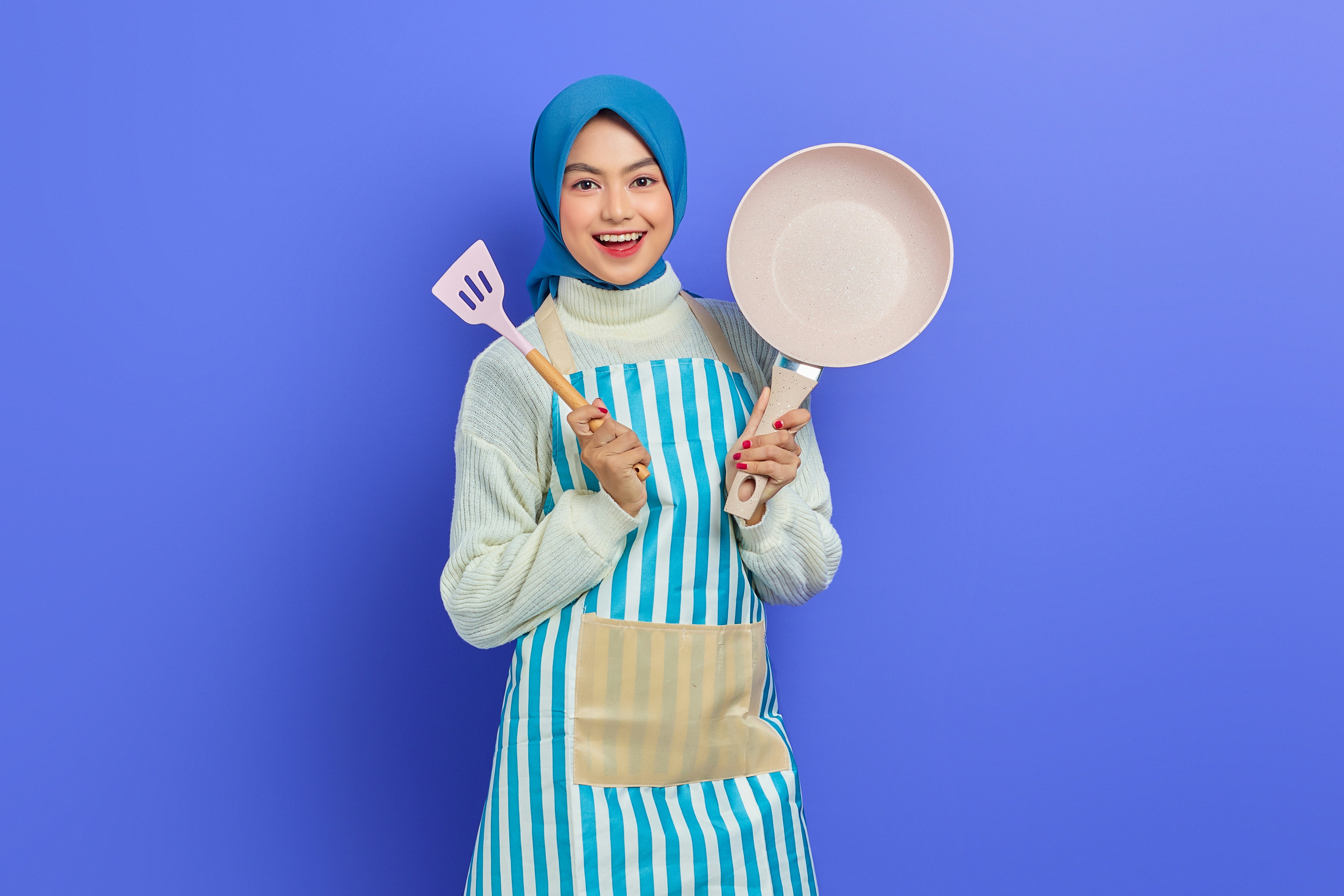 Smiling woman in a blue hijab and striped apron holds a frying pan and spatula against a vibrant purple backdrop.