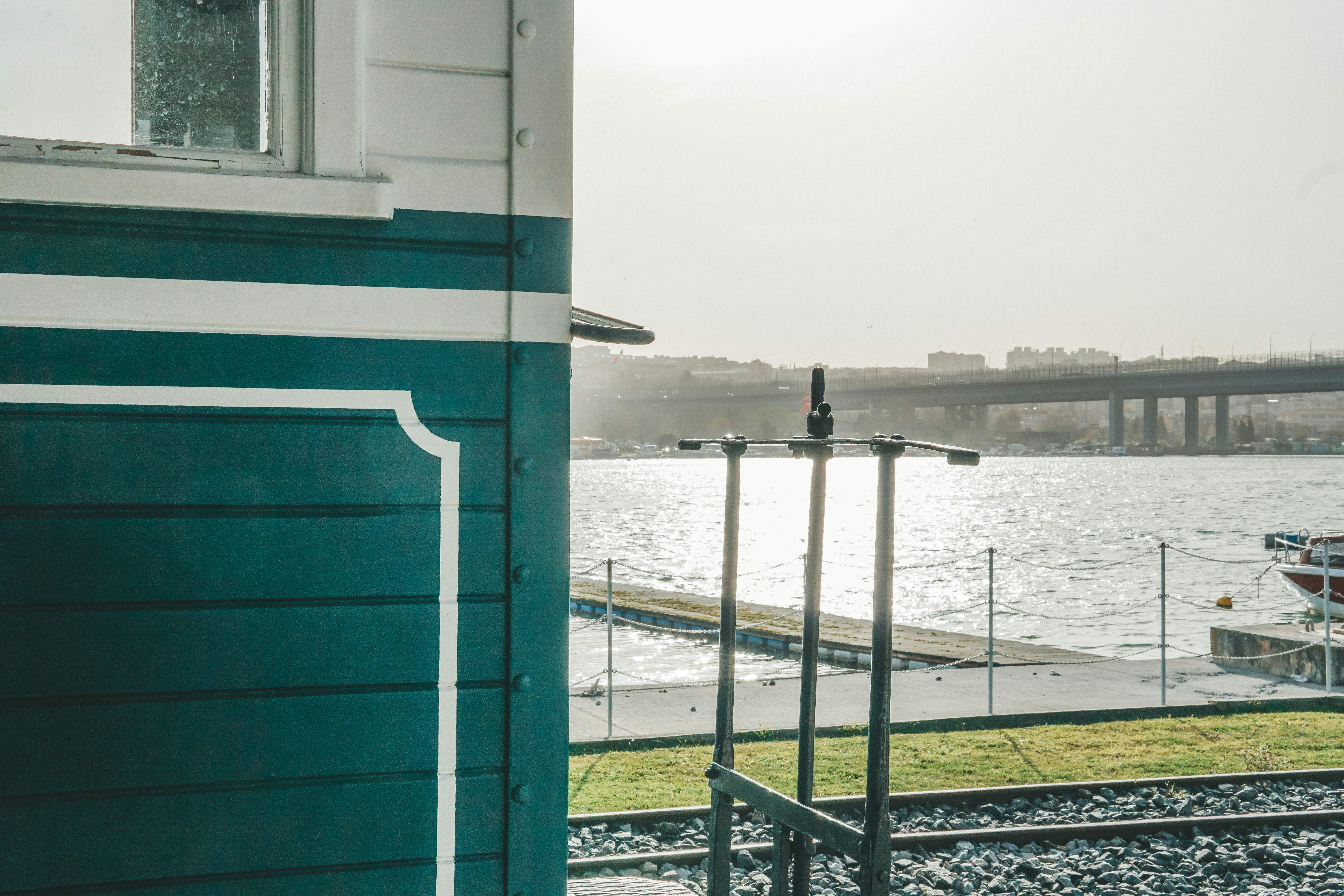 Teal wooden structure framing a serene harbor view with a distant bridge and boats. Soft sunlight reflects off the water's surface.