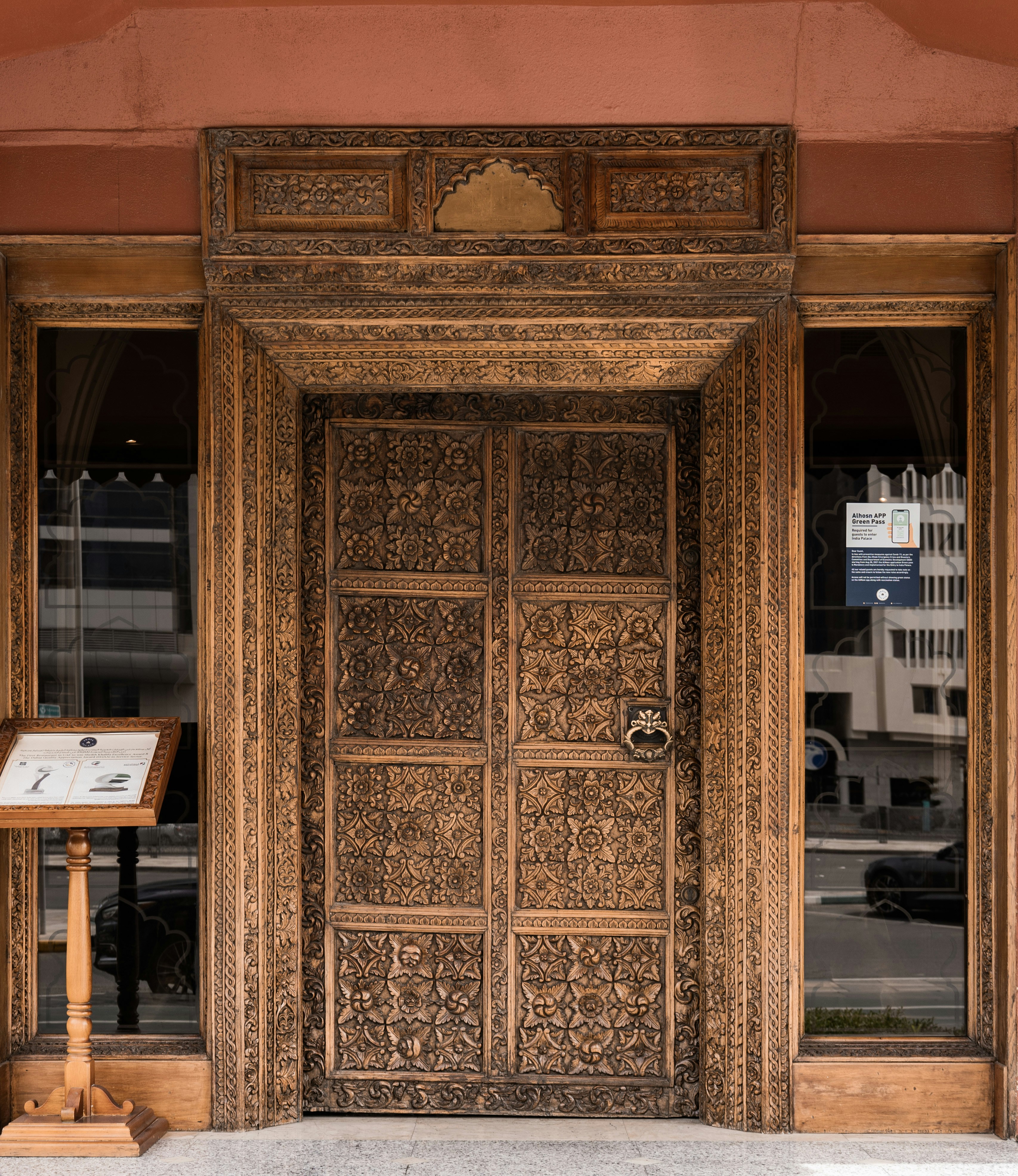 Ornately carved wooden door framed by elegant architecture, showcasing intricate patterns and details. A small sign and display stand complement the entrance.