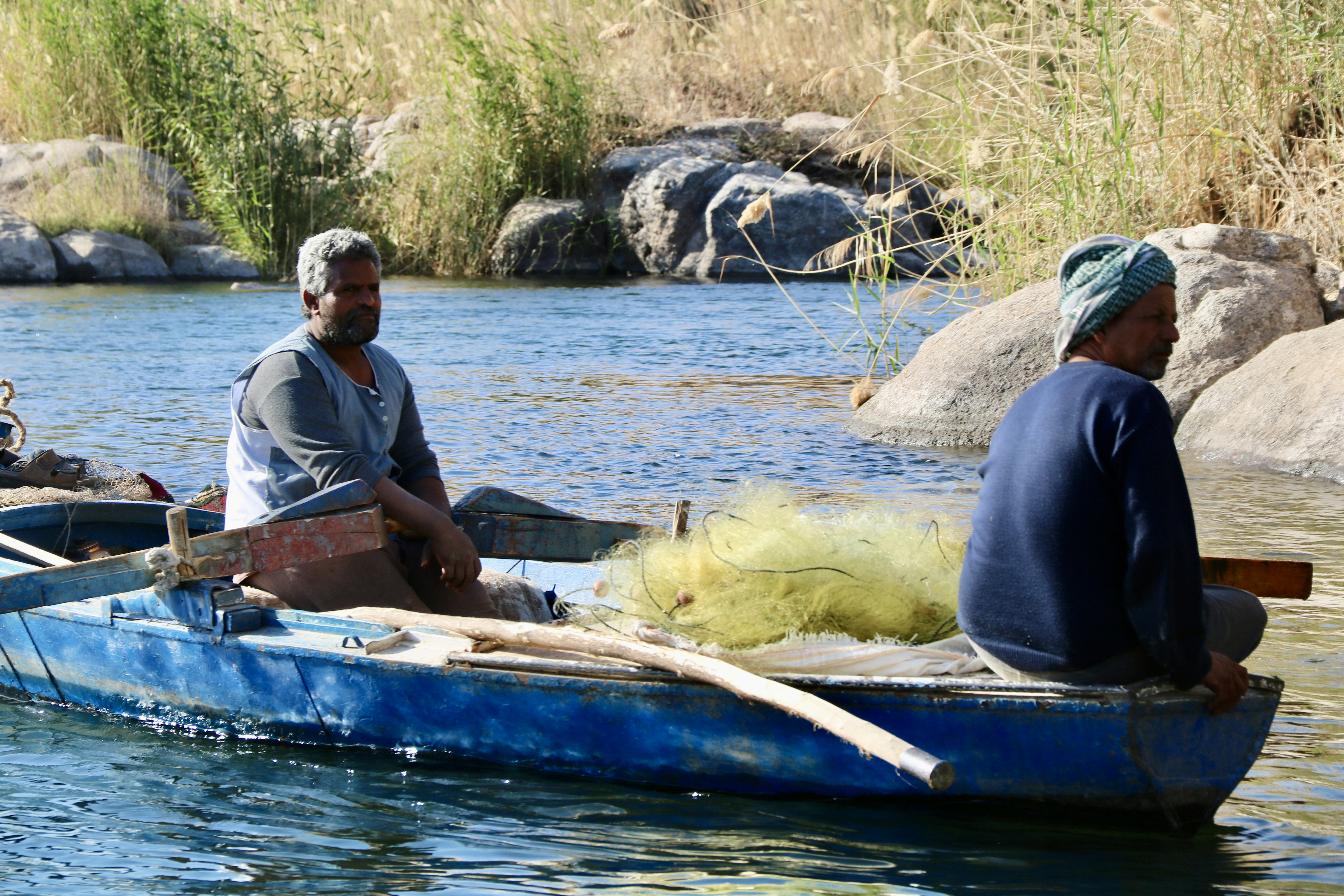 Two fishermen in a blue boat navigate a serene river, surrounded by rocky banks and tall grasses.
