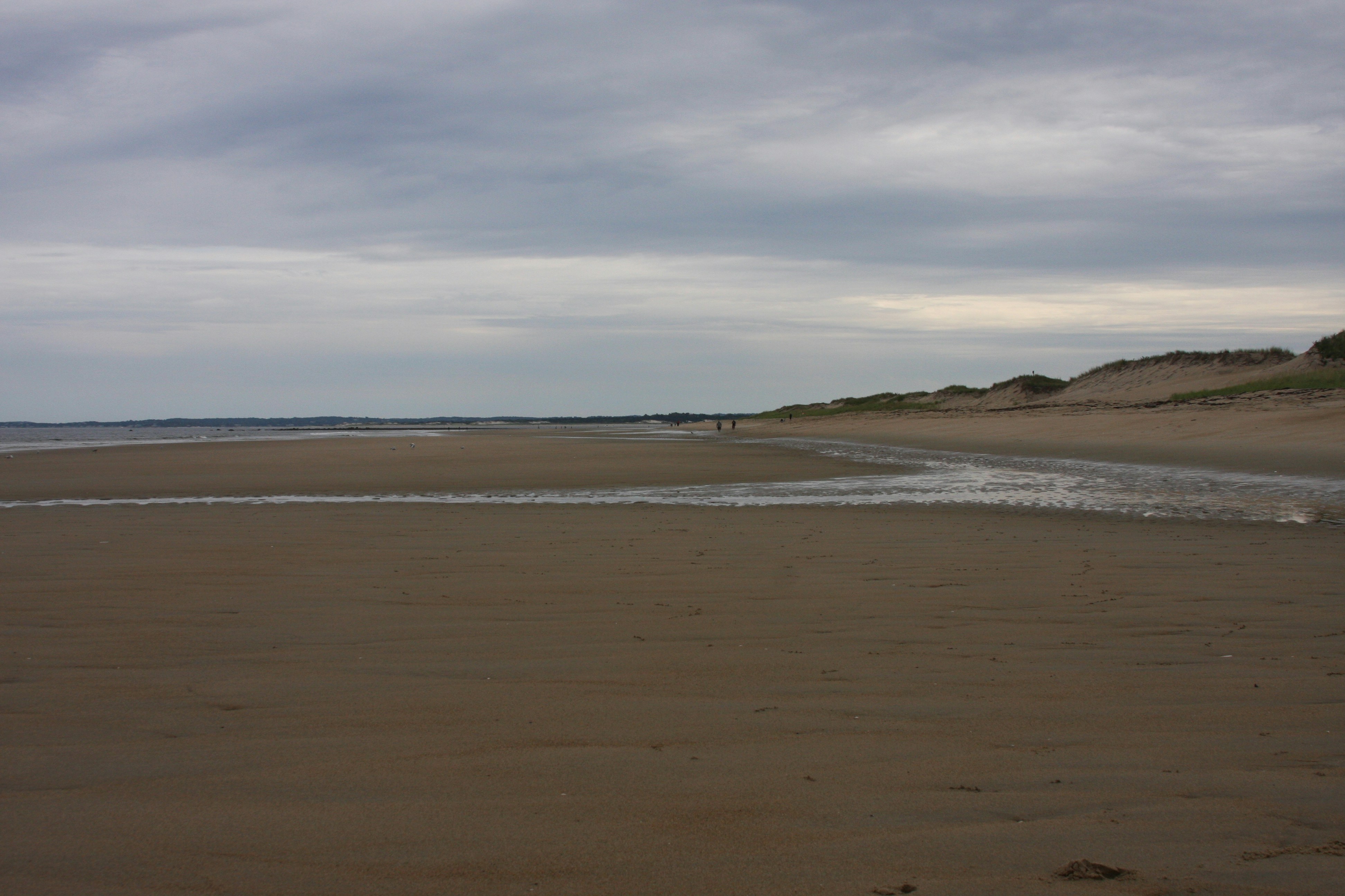 Expansive sandy beach under a cloudy sky, with a gentle tide reflecting the landscape. Distant figures stroll along the shoreline.