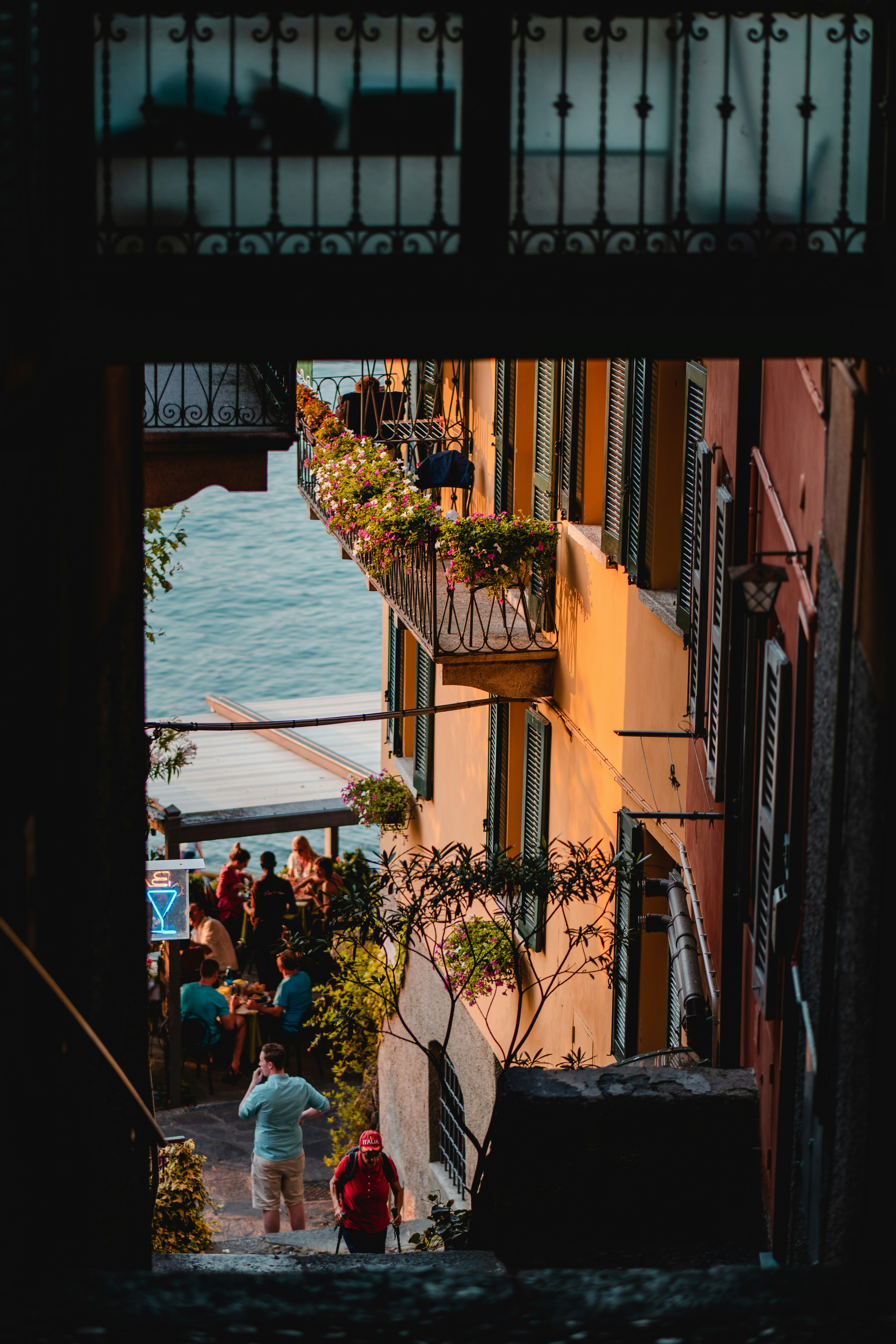 Vibrant street scene in a coastal town, showcasing colorful buildings adorned with flowers and people enjoying the ambiance near the water.