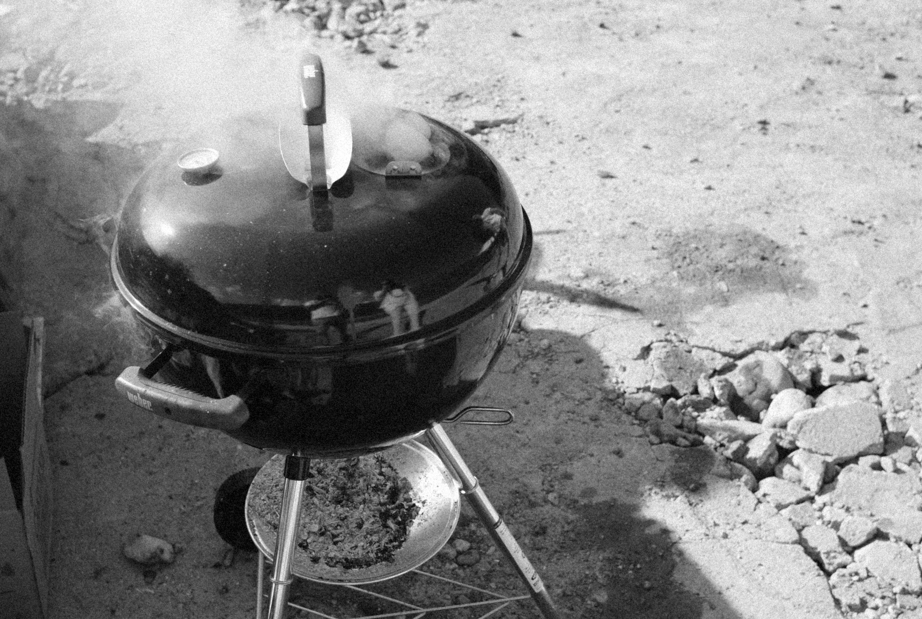 Black-and-white photograph of a round kettle charcoal grill on rocky ground, steam rising from its vents.