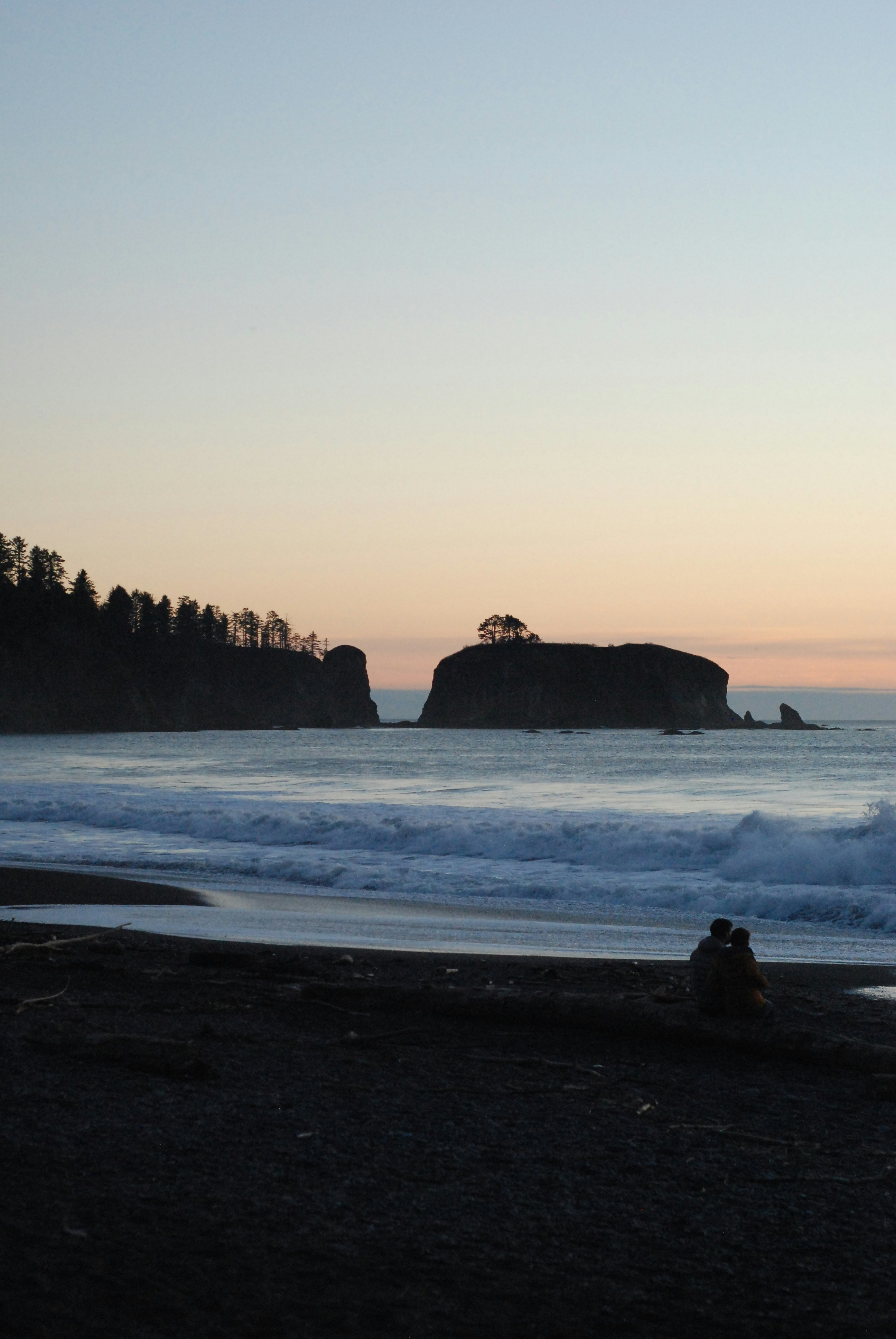 Couple seated on a beach, gazing at the ocean and distant rocky outcrops during twilight. The serene atmosphere captures a moment of connection amidst nature.