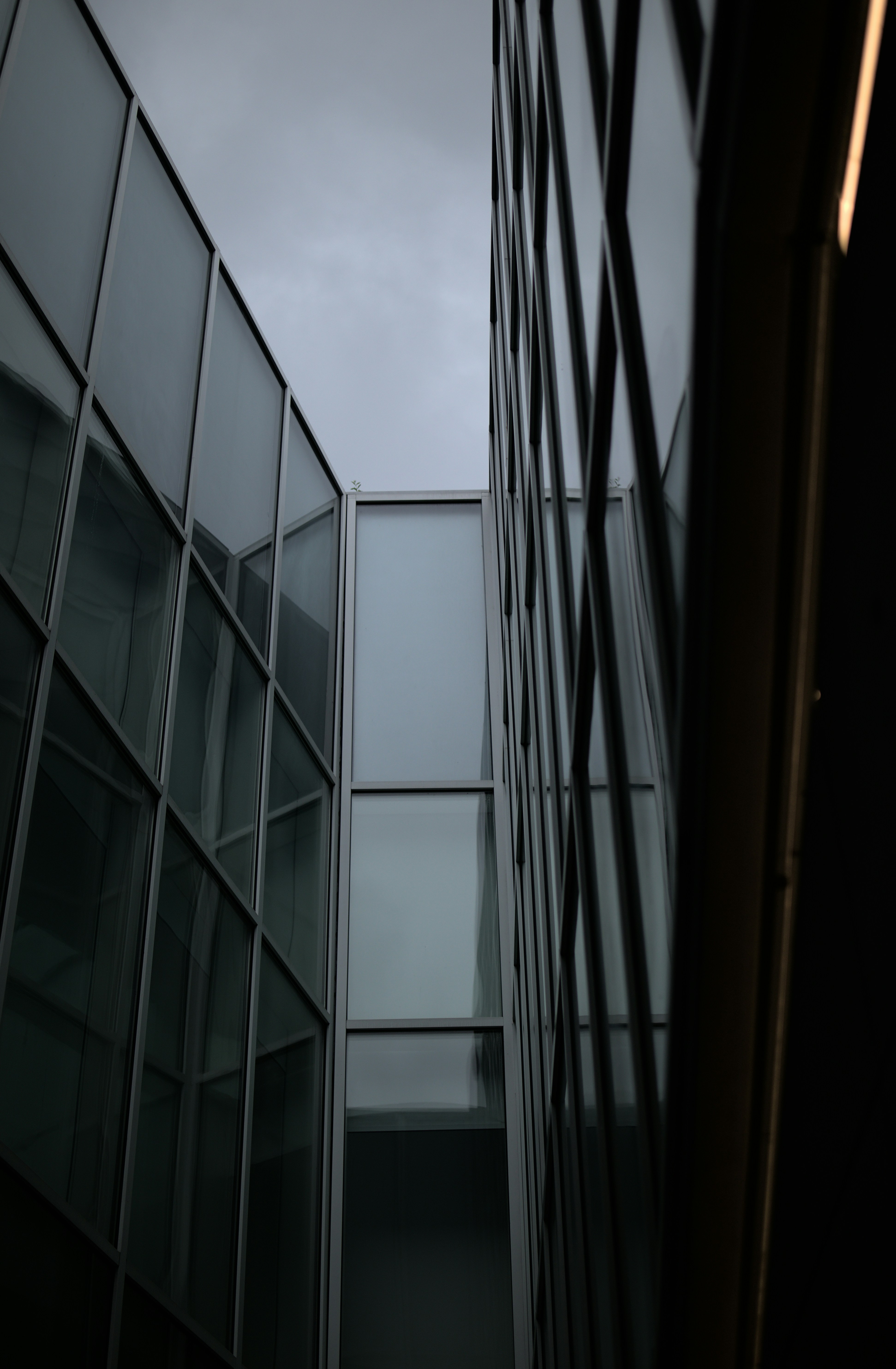 Tall glass buildings converge in a narrow alleyway, reflecting the overcast sky above. The interplay of light and shadow creates a dramatic urban scene.