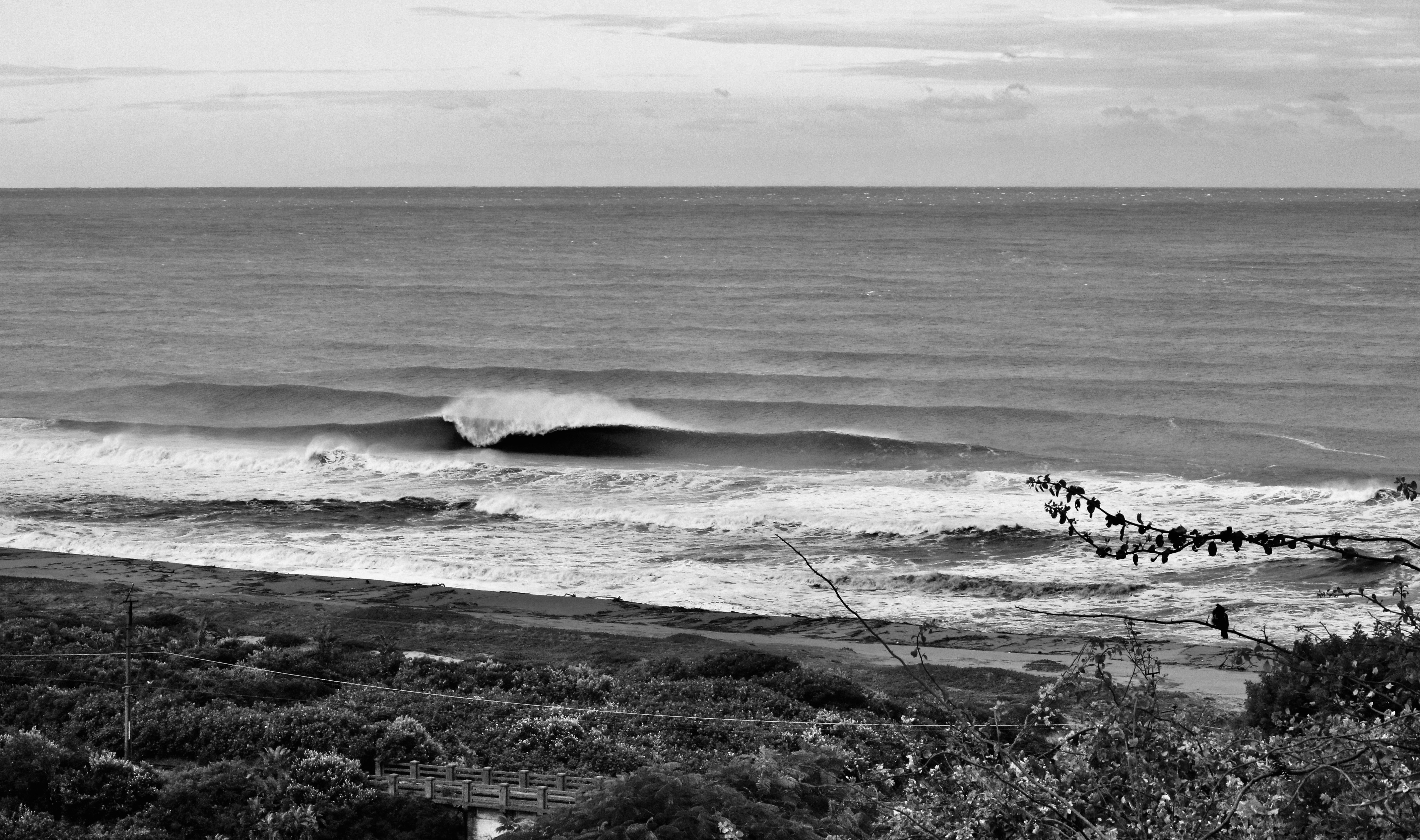 Black and white seascape depicting a large wave crashing onto a shoreline with distant clouds.