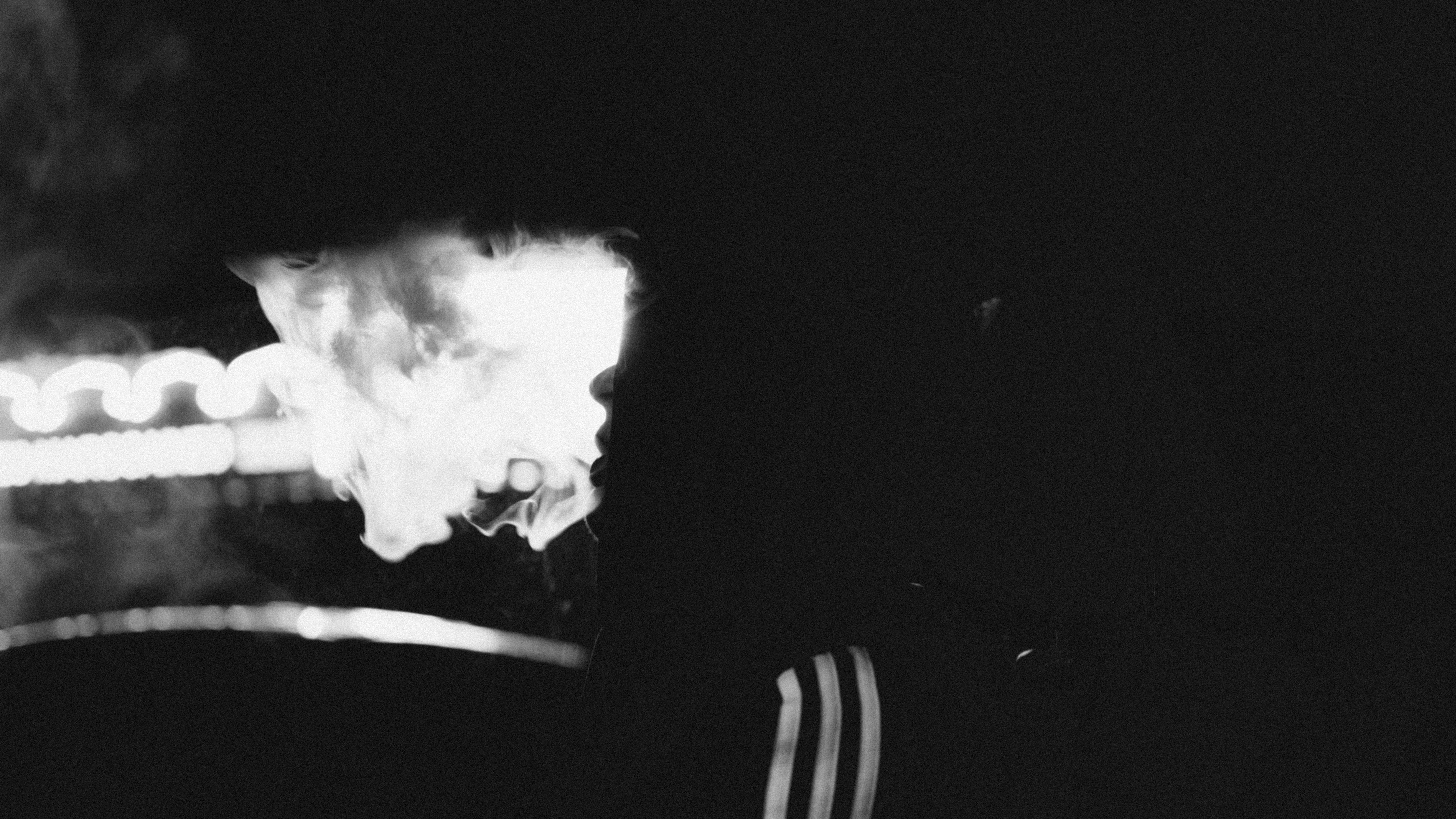 A figure shrouded in darkness exhales a plume of smoke, illuminated by subtle light in the background. The contrast between light and shadow creates an intriguing atmosphere.