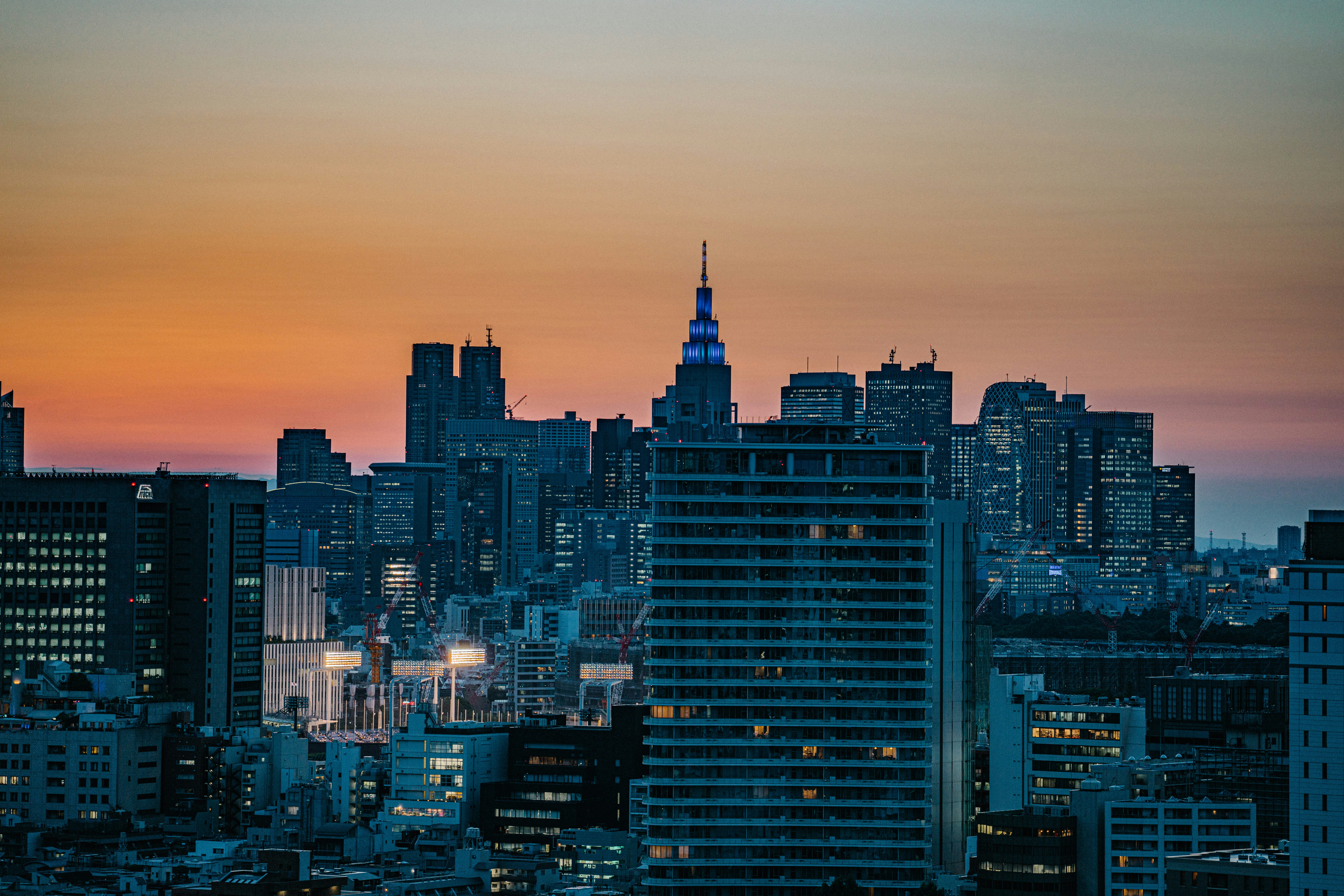 Tokyo skyline at twilight, featuring illuminated buildings and a prominent tower topped with blue lights. The gradient sky transitions from orange to deep blue.