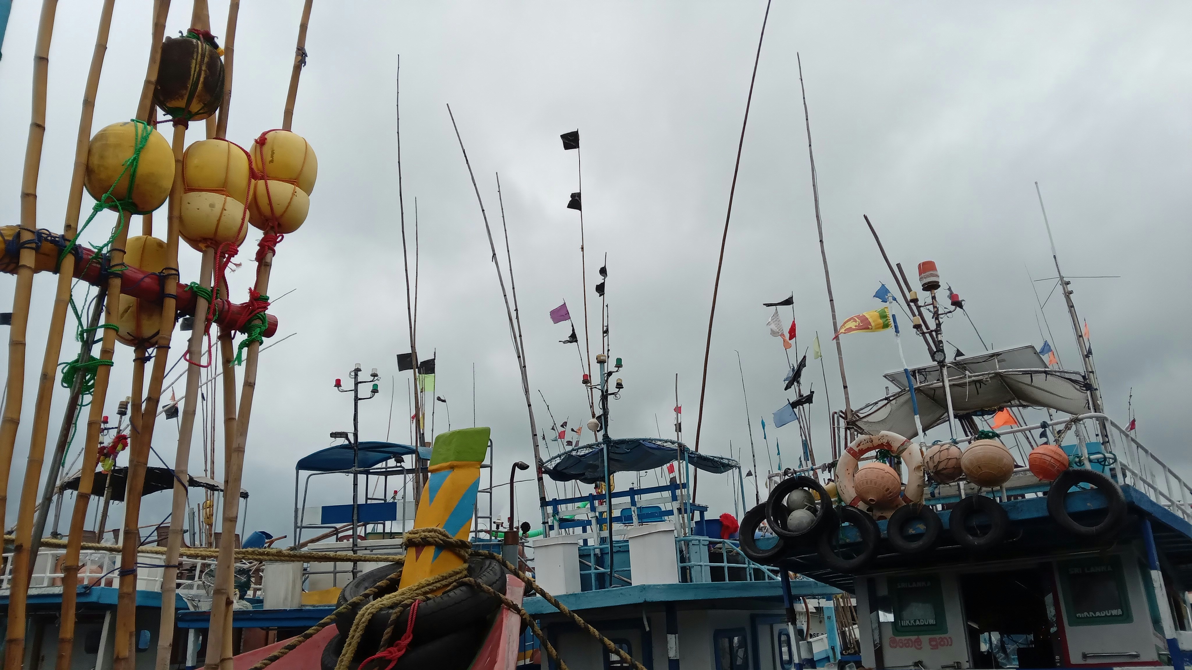 Colorful fishing gear and boats adorned with flags fill a bustling harbor under a cloudy sky.