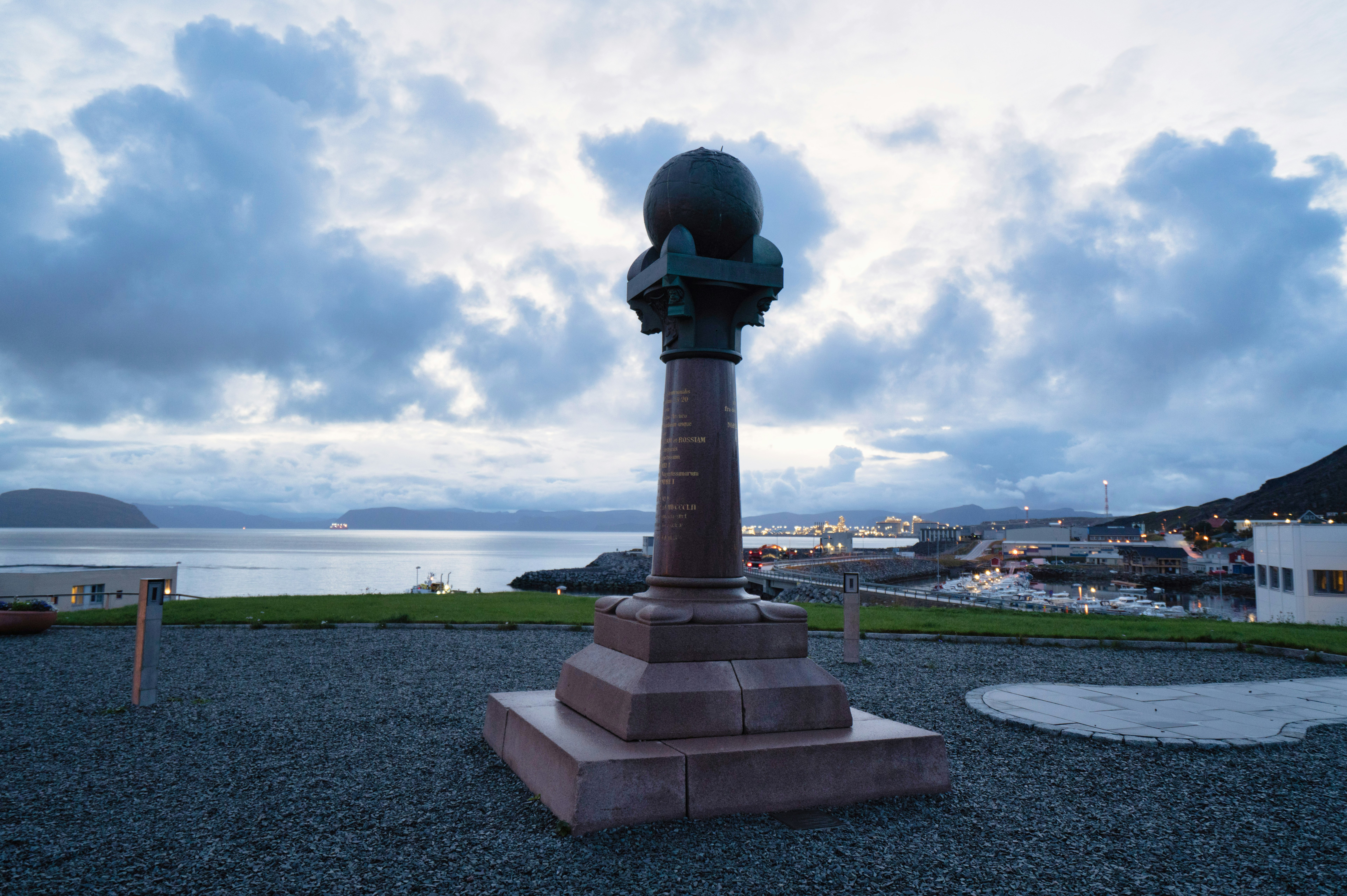 Meridian Column against a dramatic, cloud-filled sky by a calm waterfront.