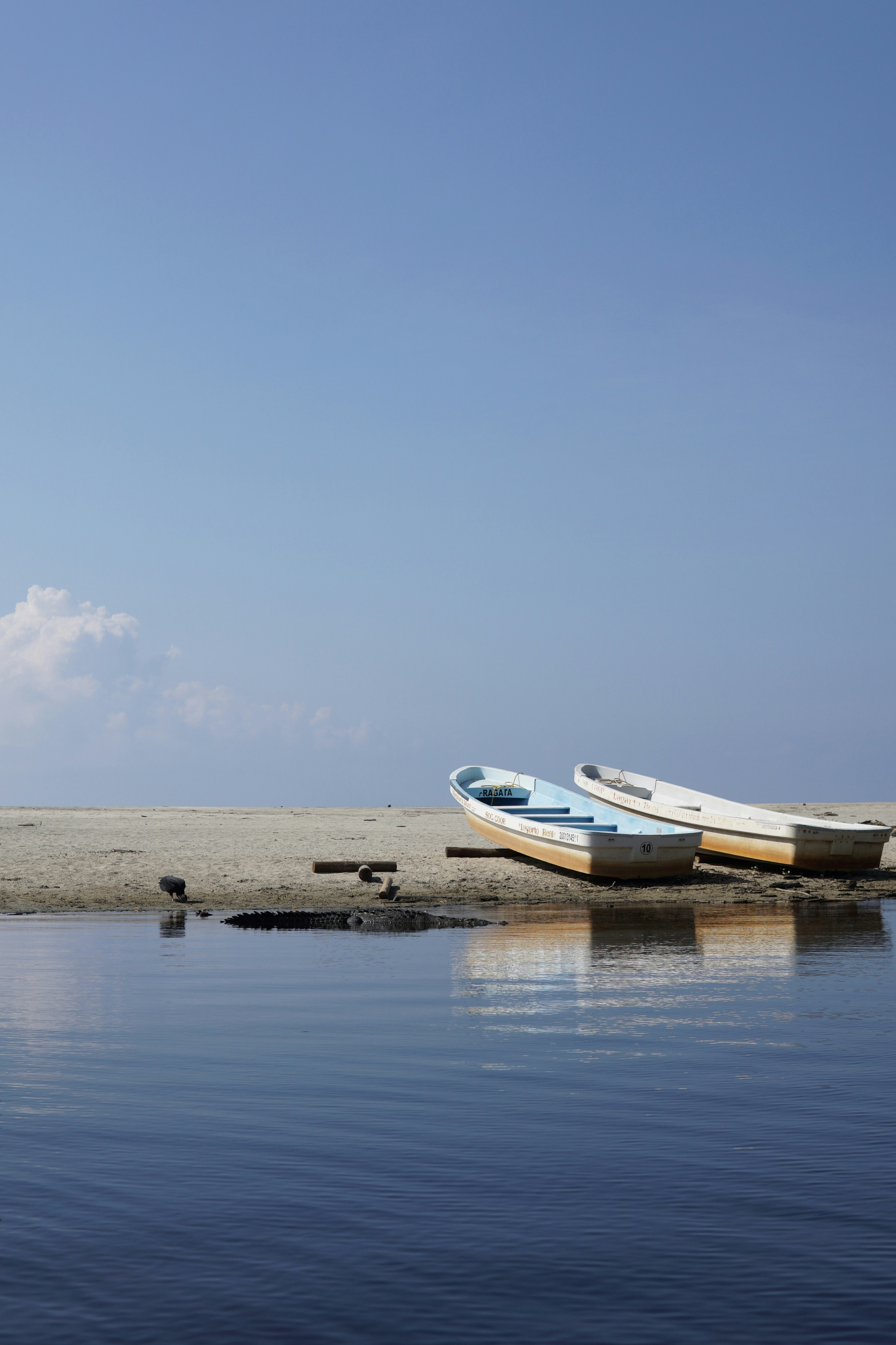 Two boats resting on a sandy beach under a clear blue sky, reflecting softly in the calm water. The tranquil setting invites a sense of peace.