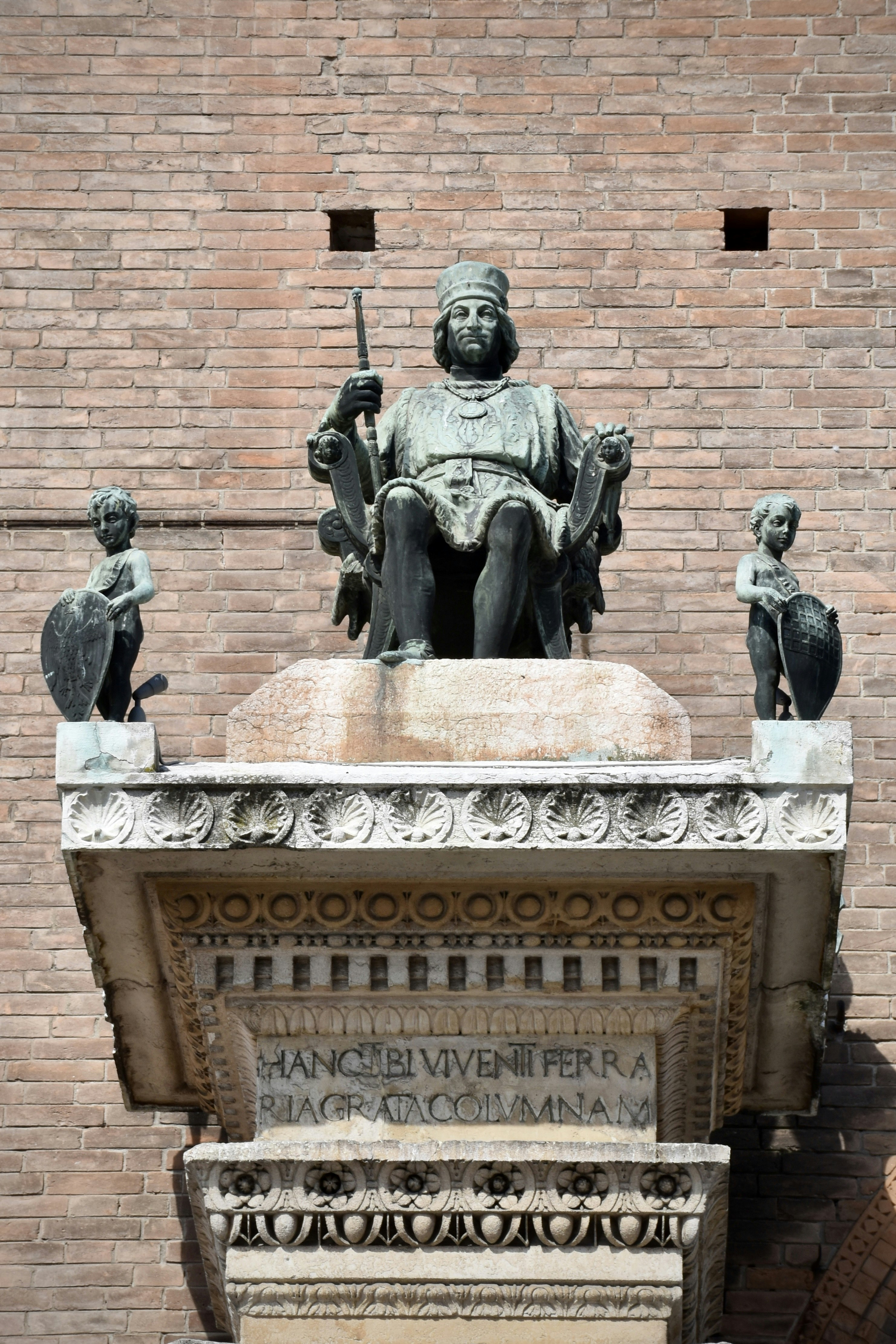 Bronze statue of a regal figure seated on a throne, flanked by two cherubs holding shields, set against a textured brick wall.