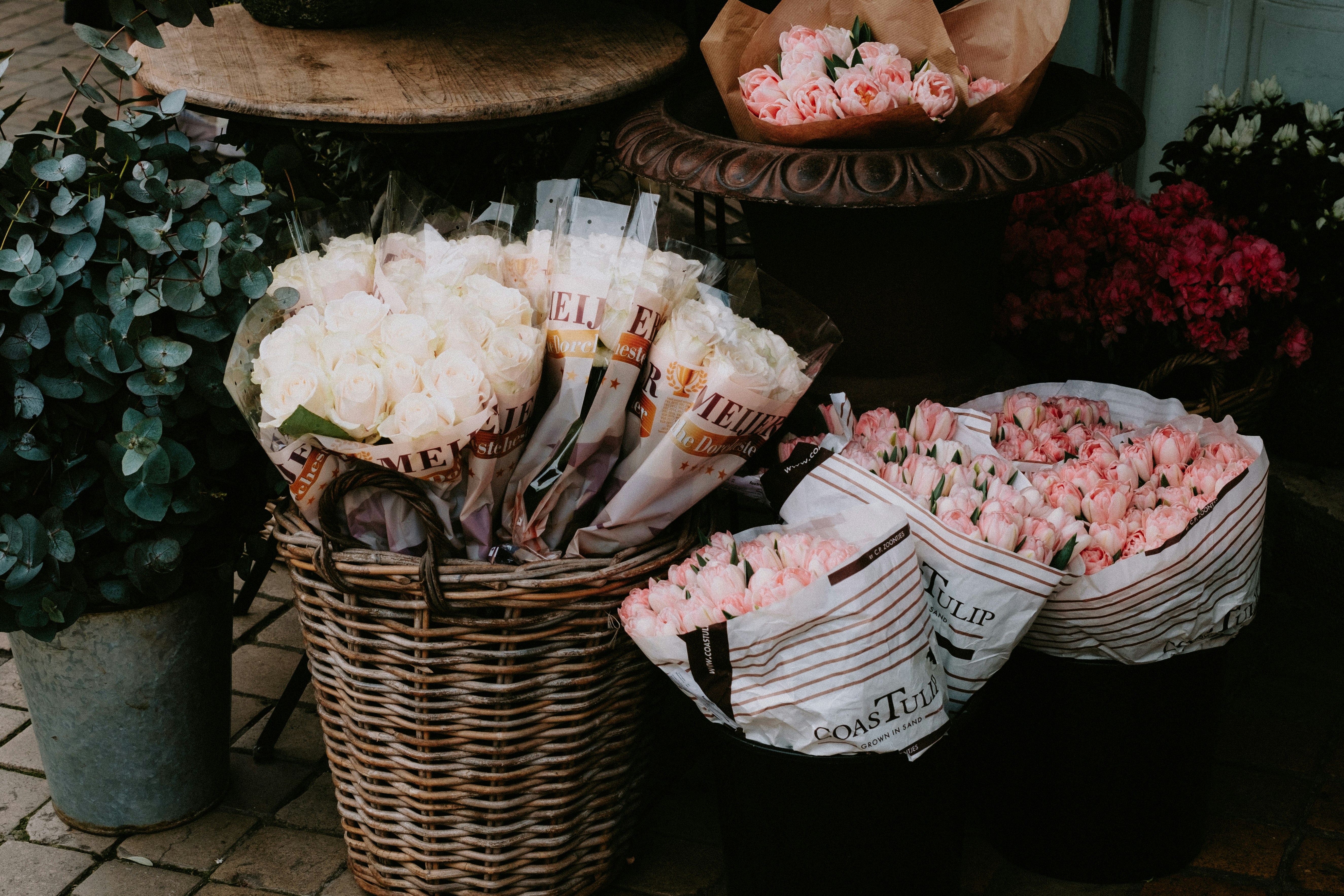 A charming flower shop display featuring an array of pink and white rose bouquets arranged in wicker baskets and bags, surrounded by lush greenery.
