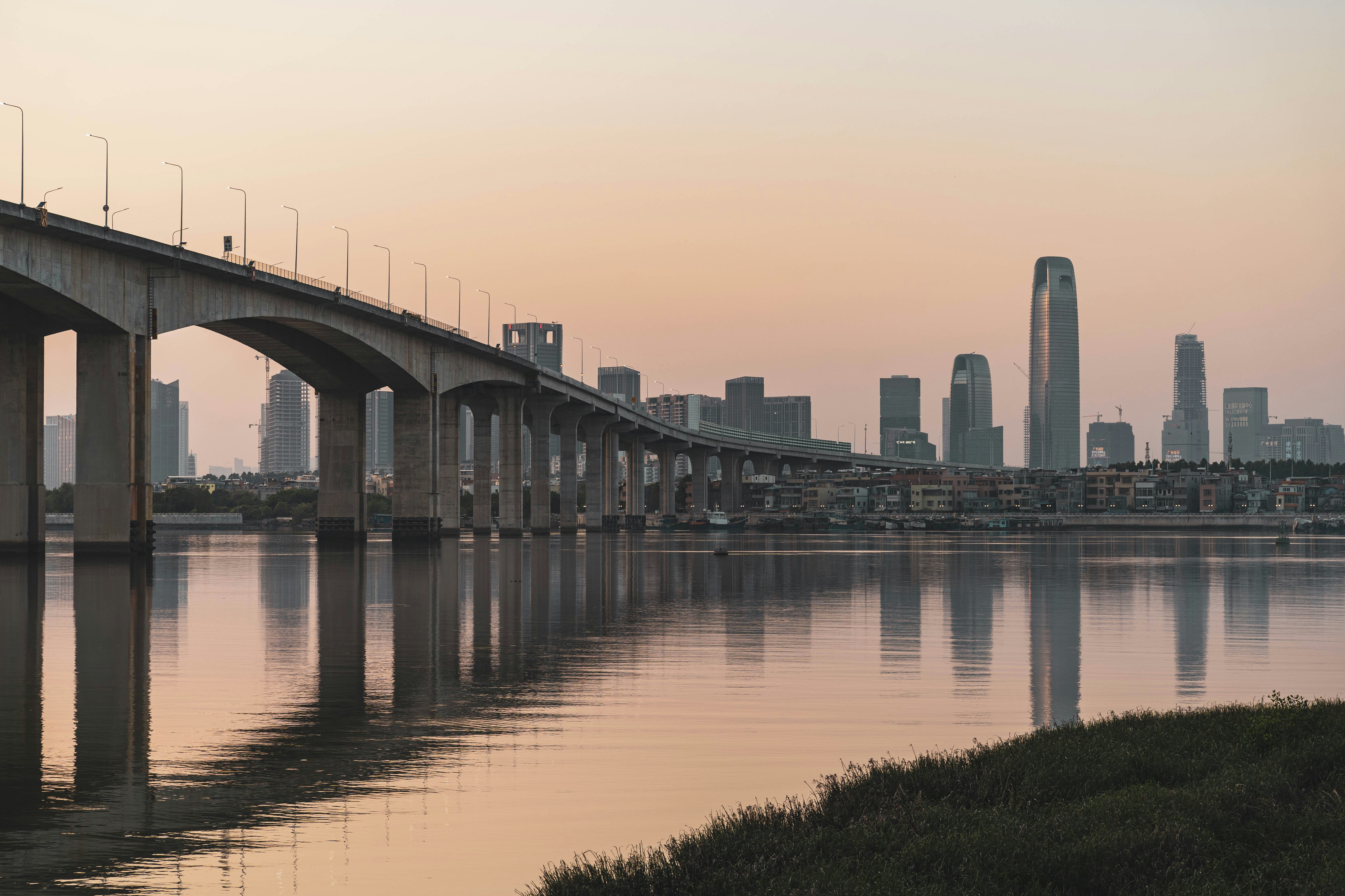 Bridge arches gracefully over a calm river at dusk with a city skyline in the background.